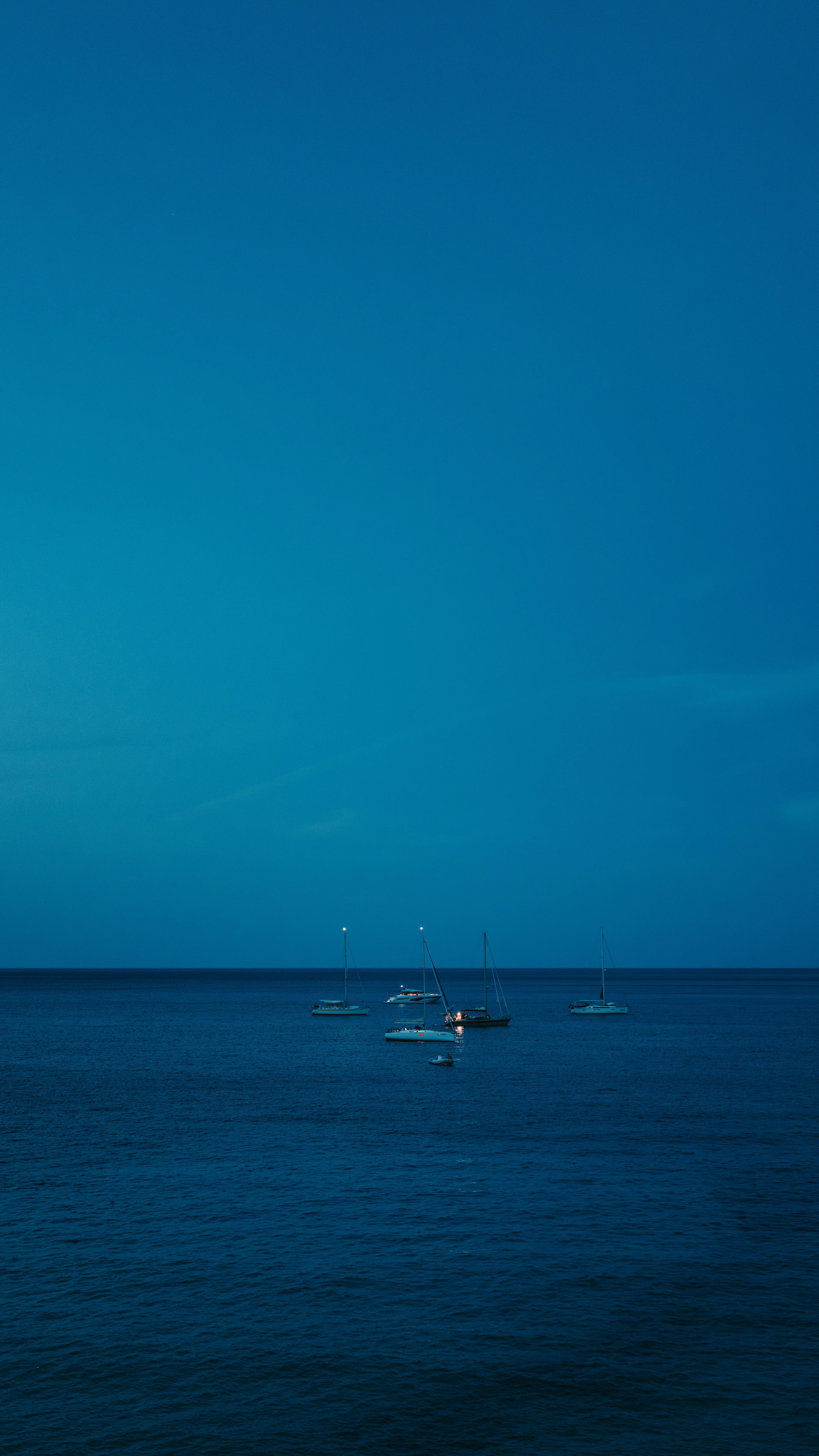 Calm sea with a small fleet of sailboats near the horizon under a gradient blue sky.