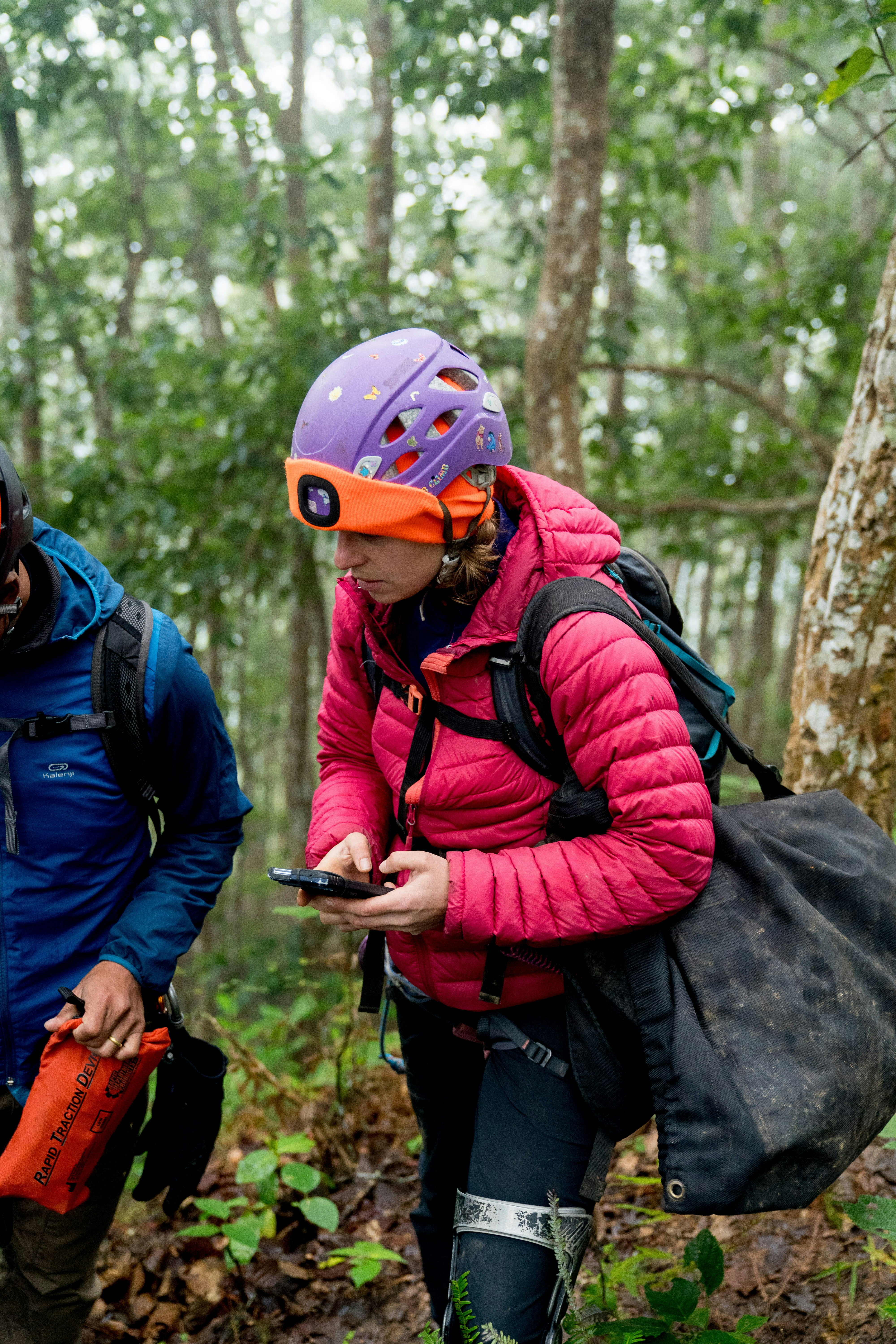 Trail Vigil: A Pink-Jacket Climber Checks her Device under a Dense Green Canopy