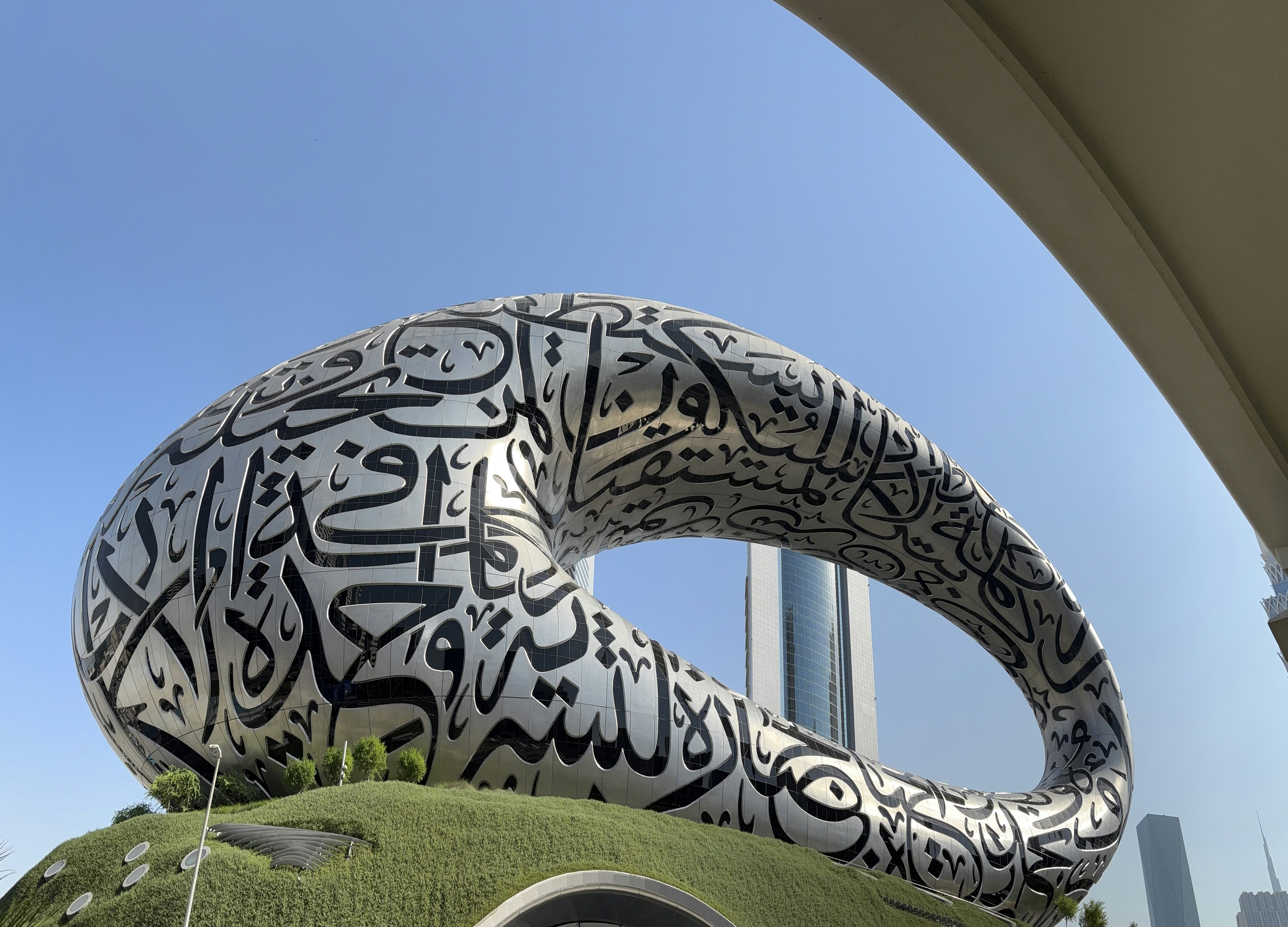 A futuristic sculpture adorned with intricate Arabic calligraphy, set against a clear blue sky. The structure blends art and architecture seamlessly.