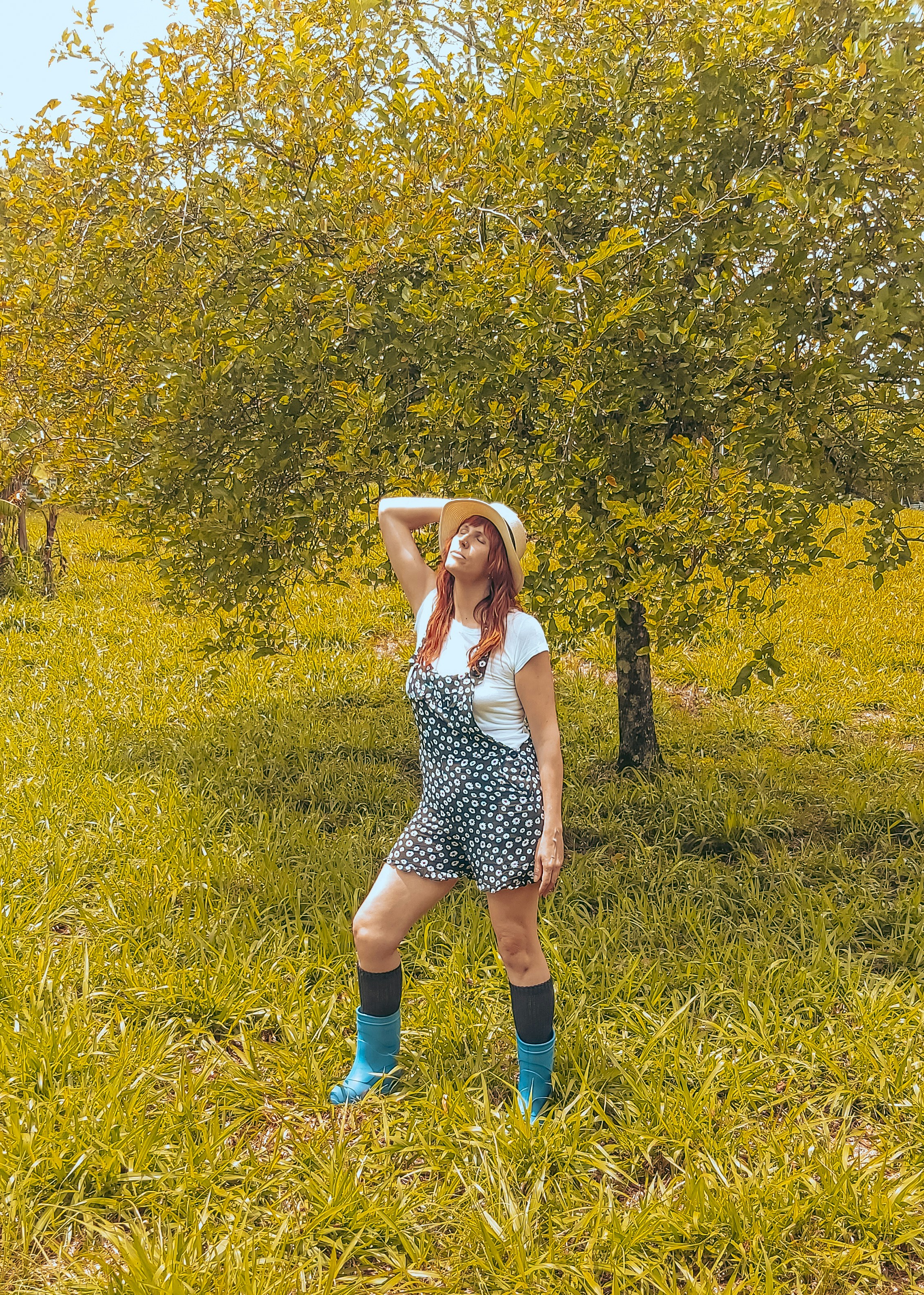 A woman poses in a sunlit field of tall yellow grass with a tree behind, wearing a hat, white top, and a floral romper.