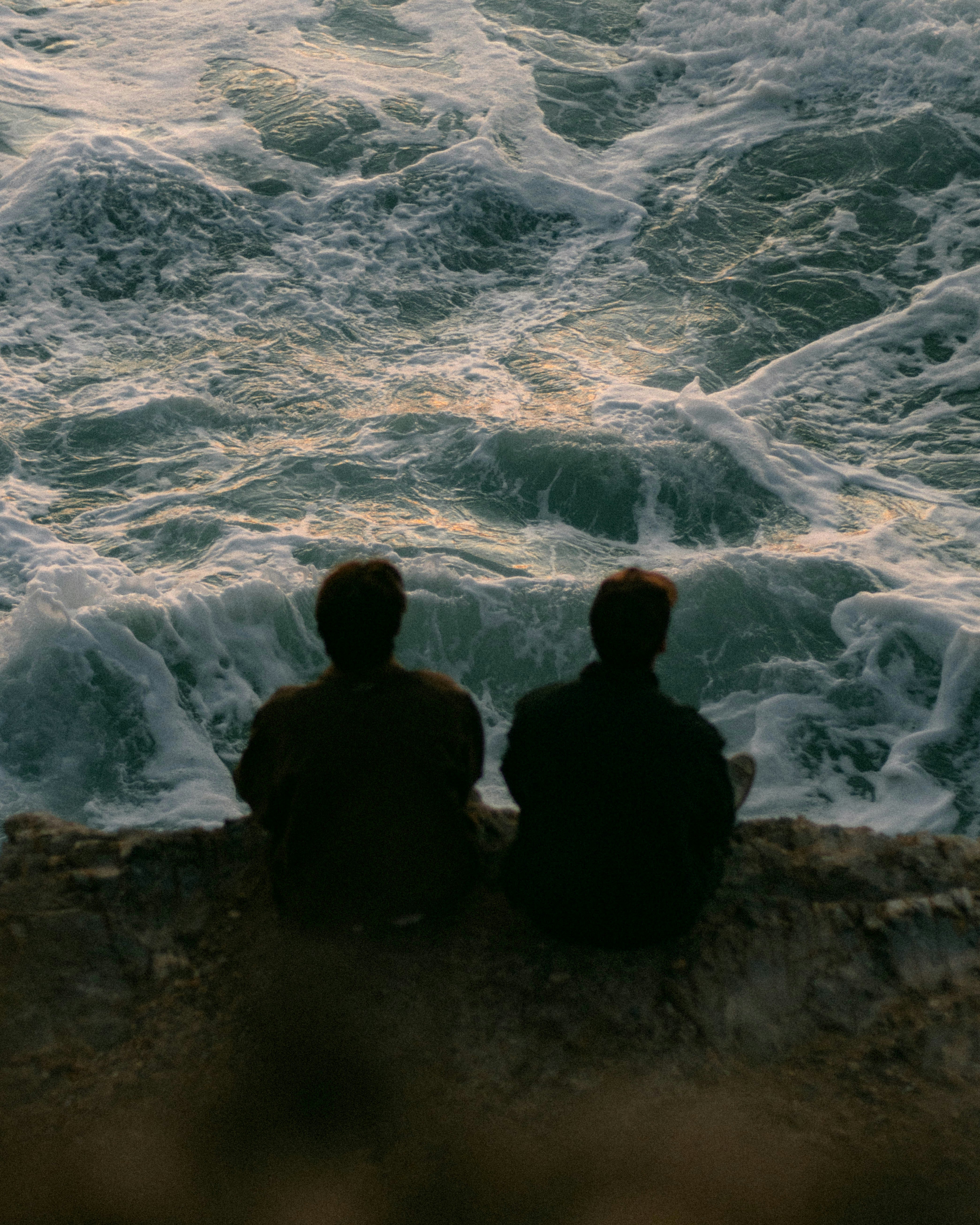 Two silhouetted figures sit on a cliff, overlooking churning ocean waves reflecting soft sunlight.
