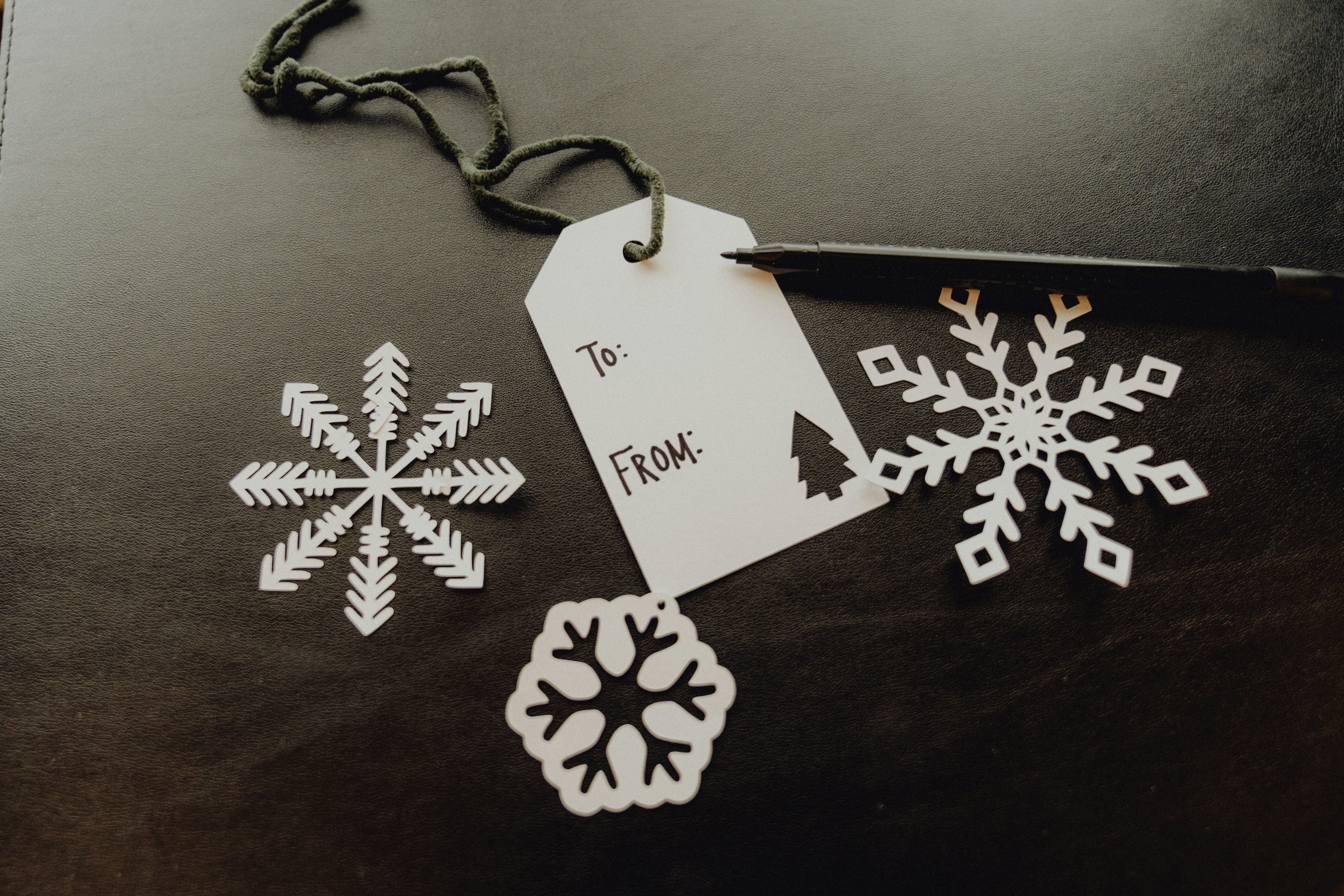 A creatively arranged gift tag with handwritten 'To:' and 'From:' alongside intricate paper snowflakes on a dark surface.