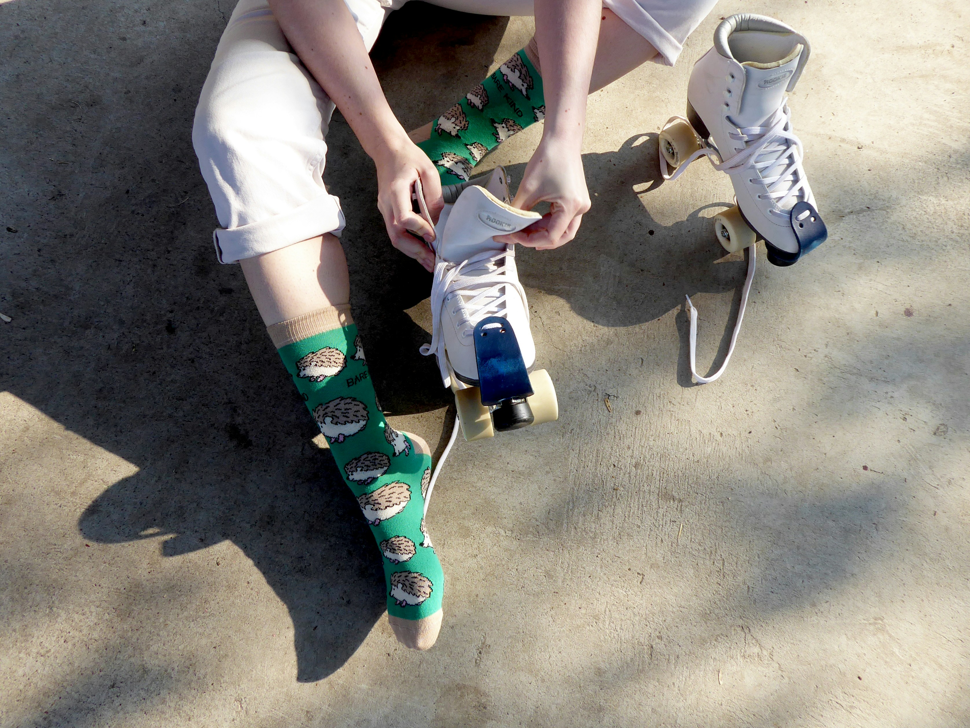 Candid photograph of someone seated on a sunlit concrete surface, tying white roller skates with panda-pattern socks visible.