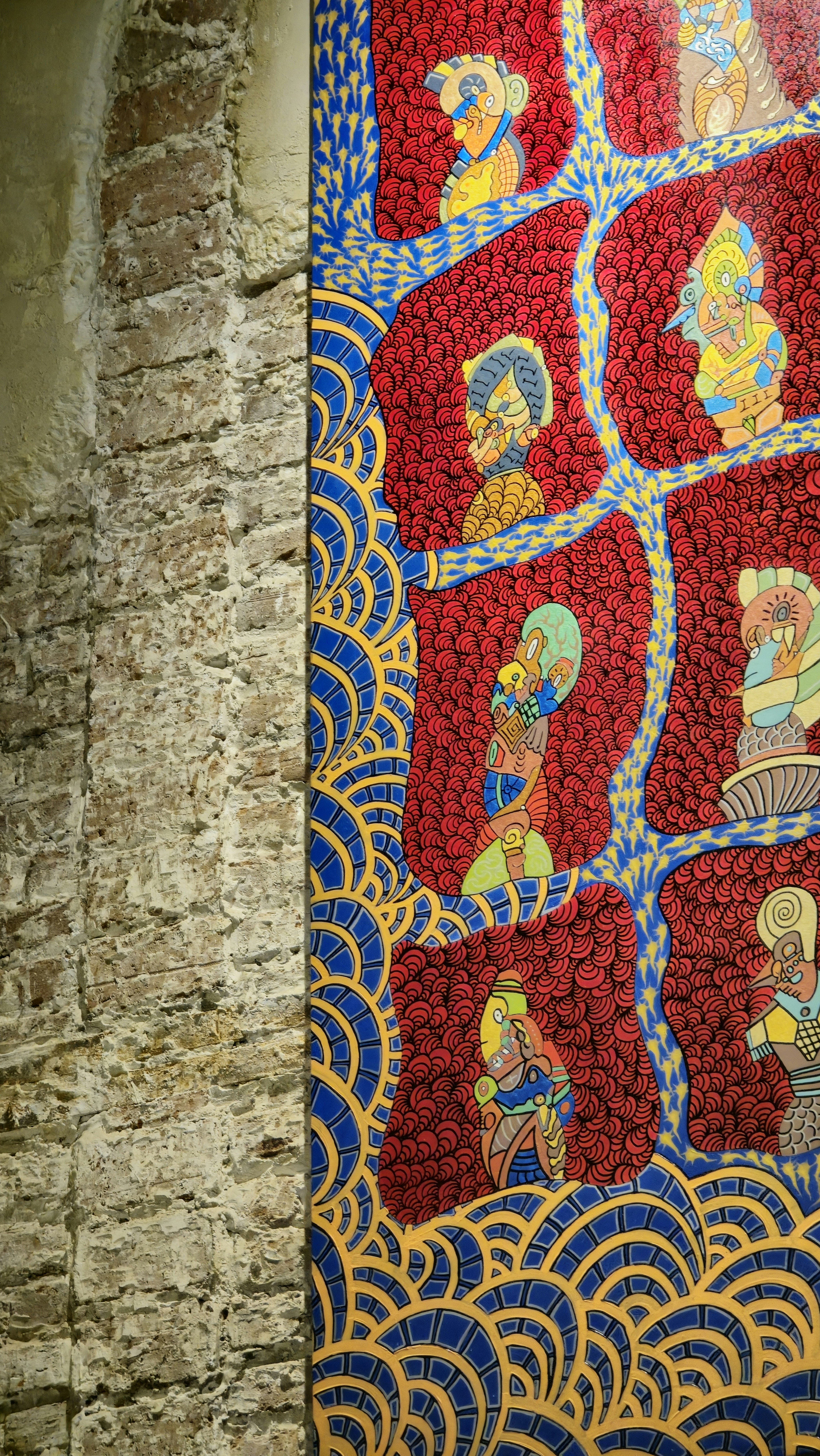 Colorful mural of stylized figures on a red field framed by blue and gold wave patterns beside a rough stone wall.