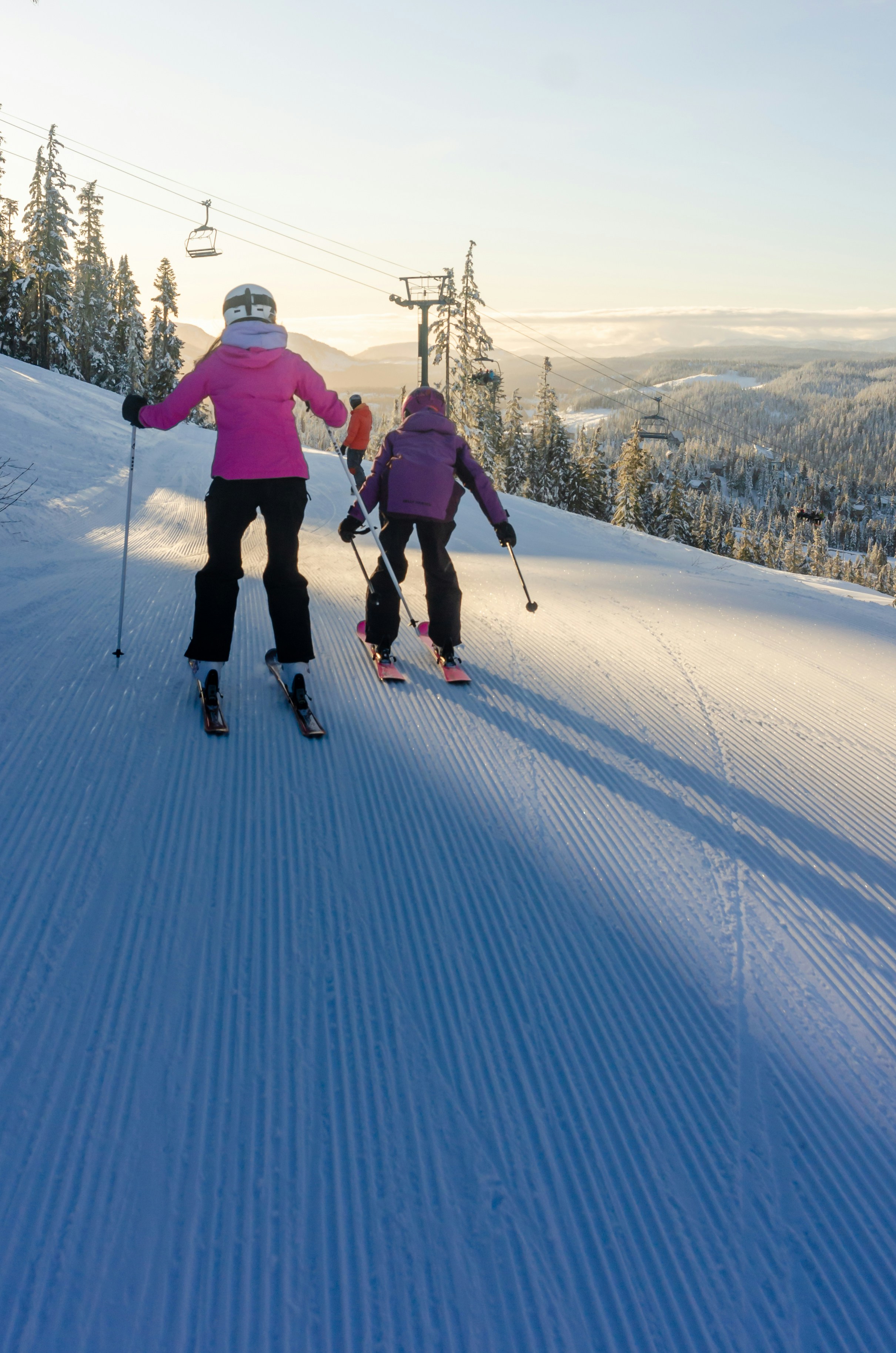 Skiers navigating freshly groomed slopes under a clear morning sky, surrounded by snow-covered trees.