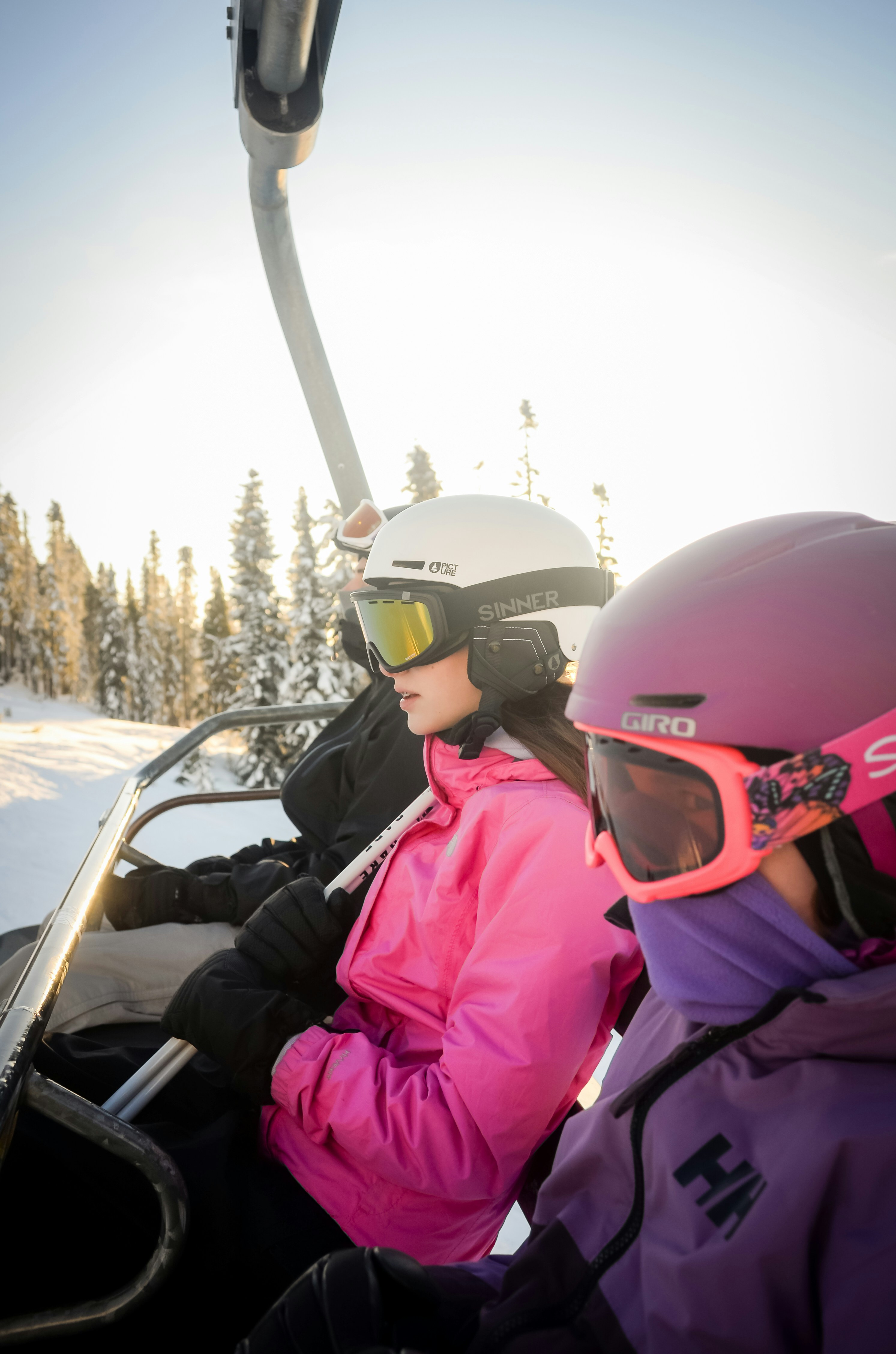 Skiers in colorful gear ride a chairlift under a bright sun, with snow-covered evergreens in the background.