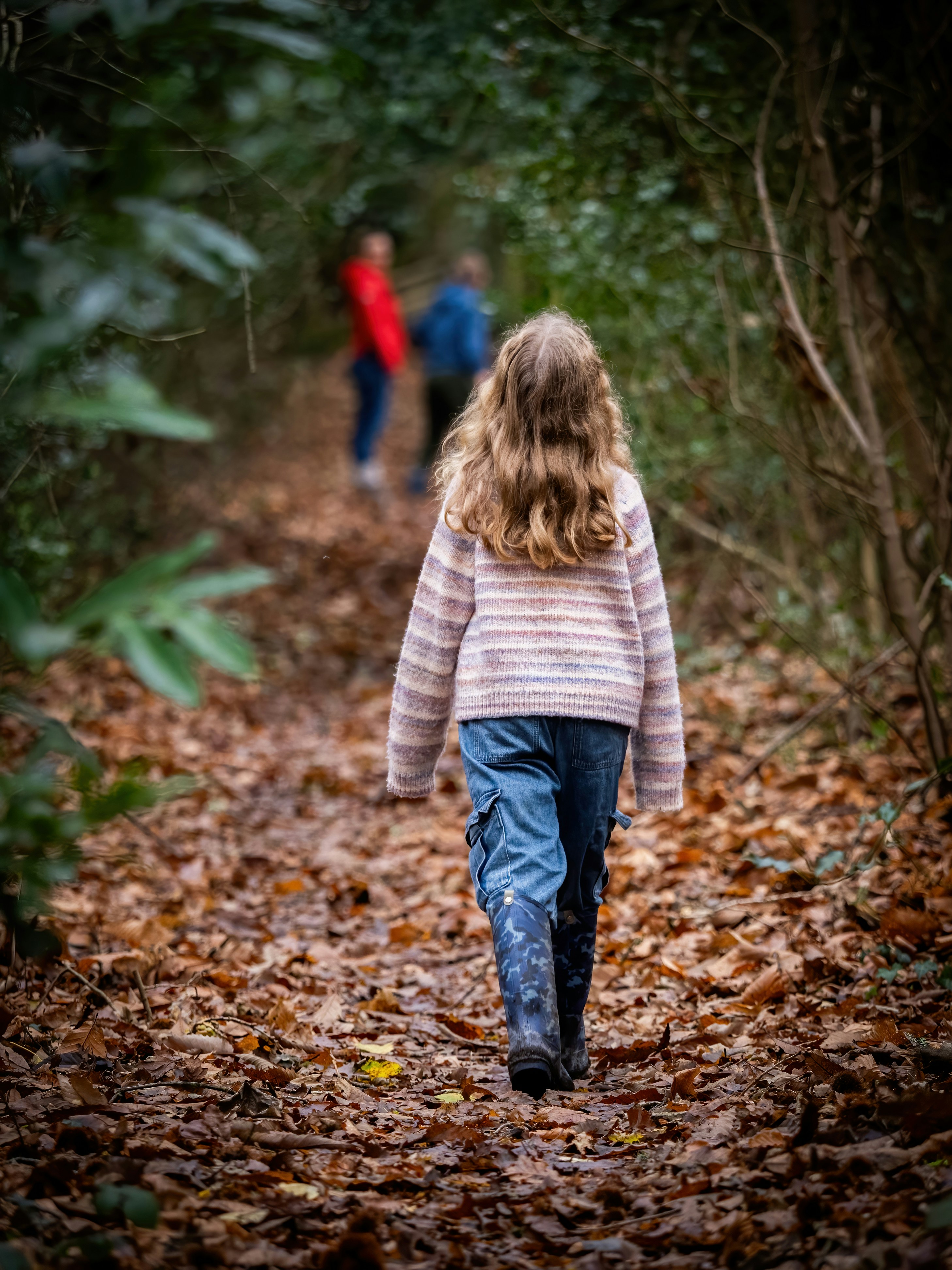 Child walking along a leaf-strewn path in a forest, surrounded by greenery and blurred figures in the background. 