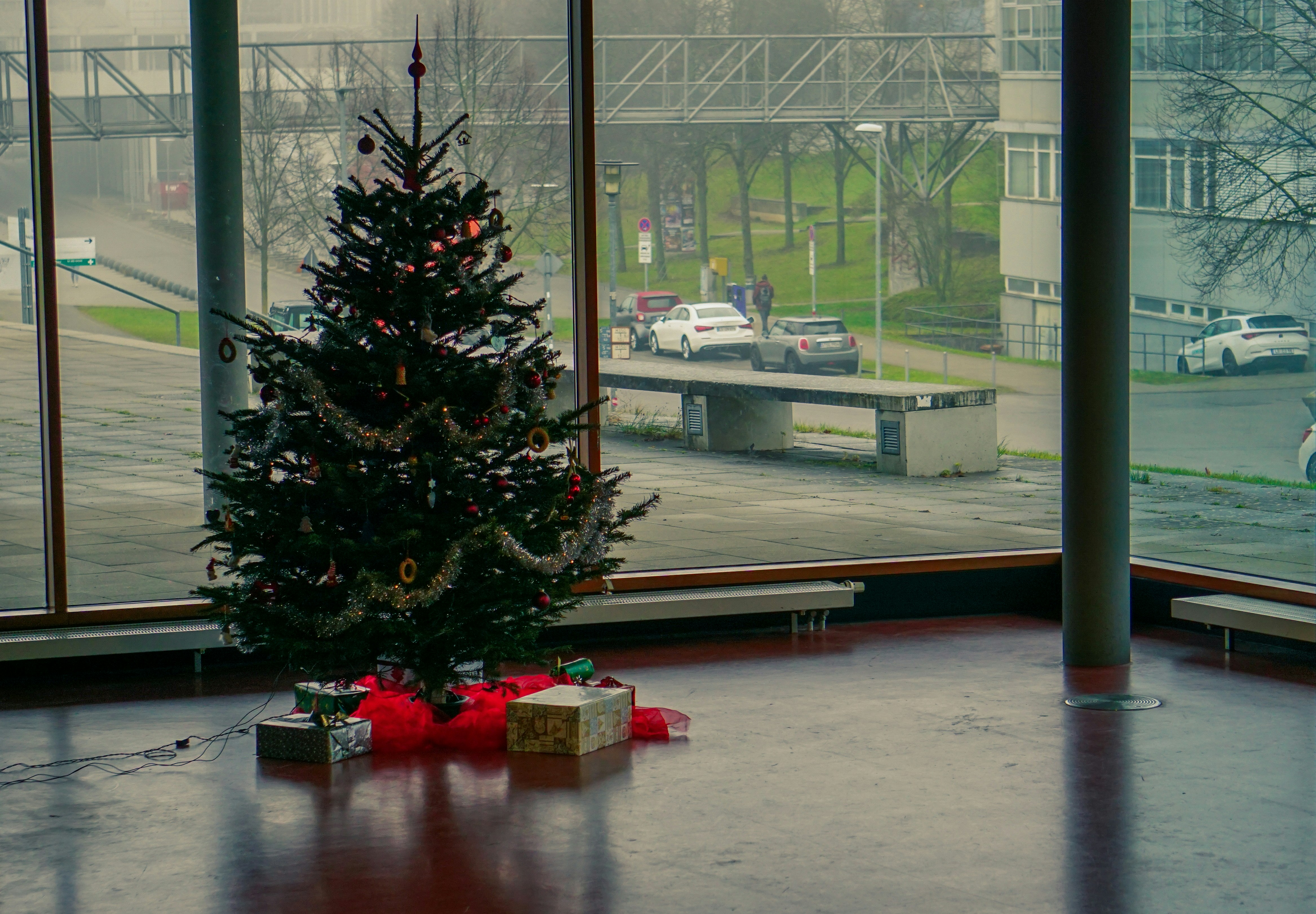 Decorated Christmas tree with gifts in a modern room with large windows overlooking an urban landscape.