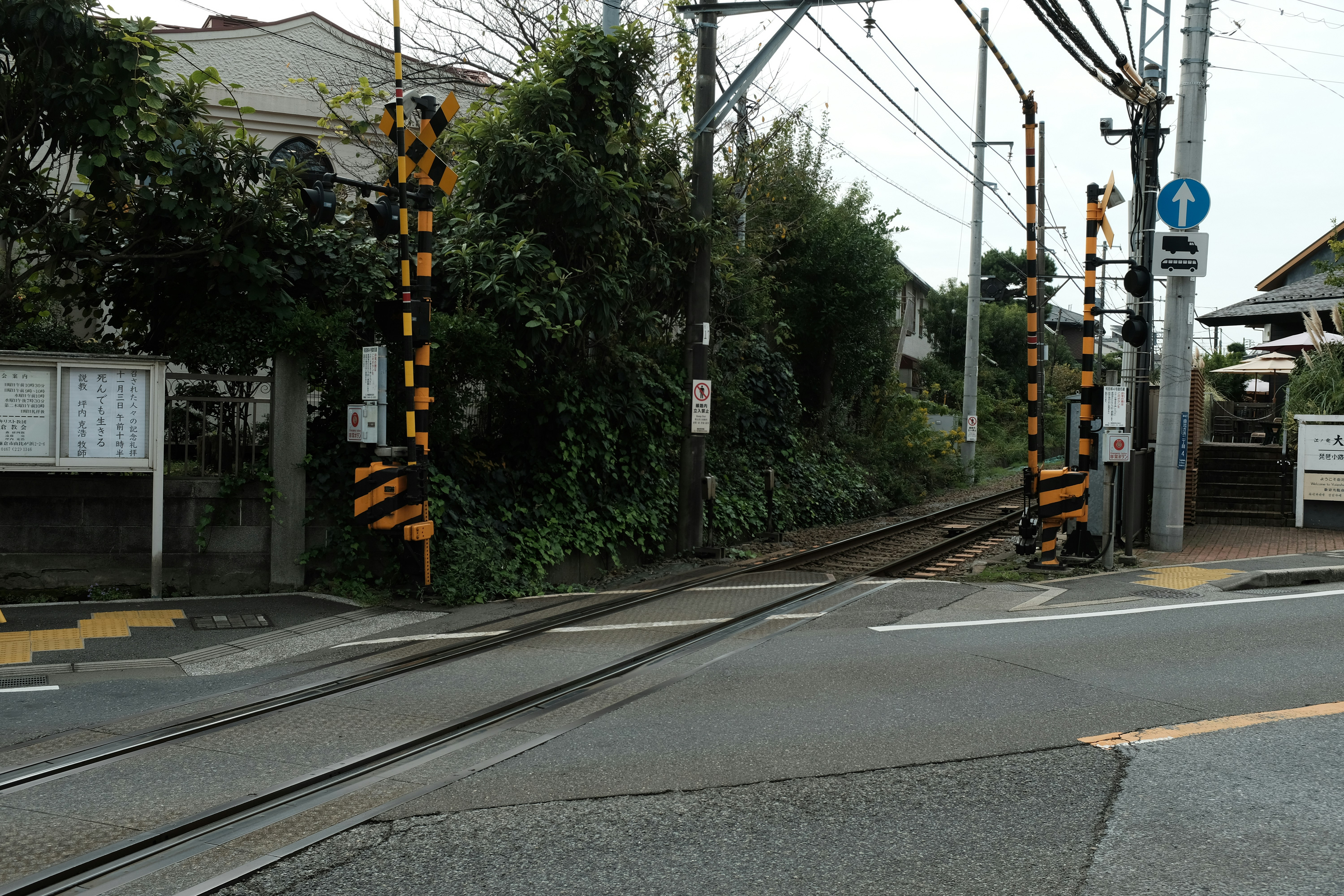 Railway crossing flanked by lush greenery and residential buildings under a cloudy sky.