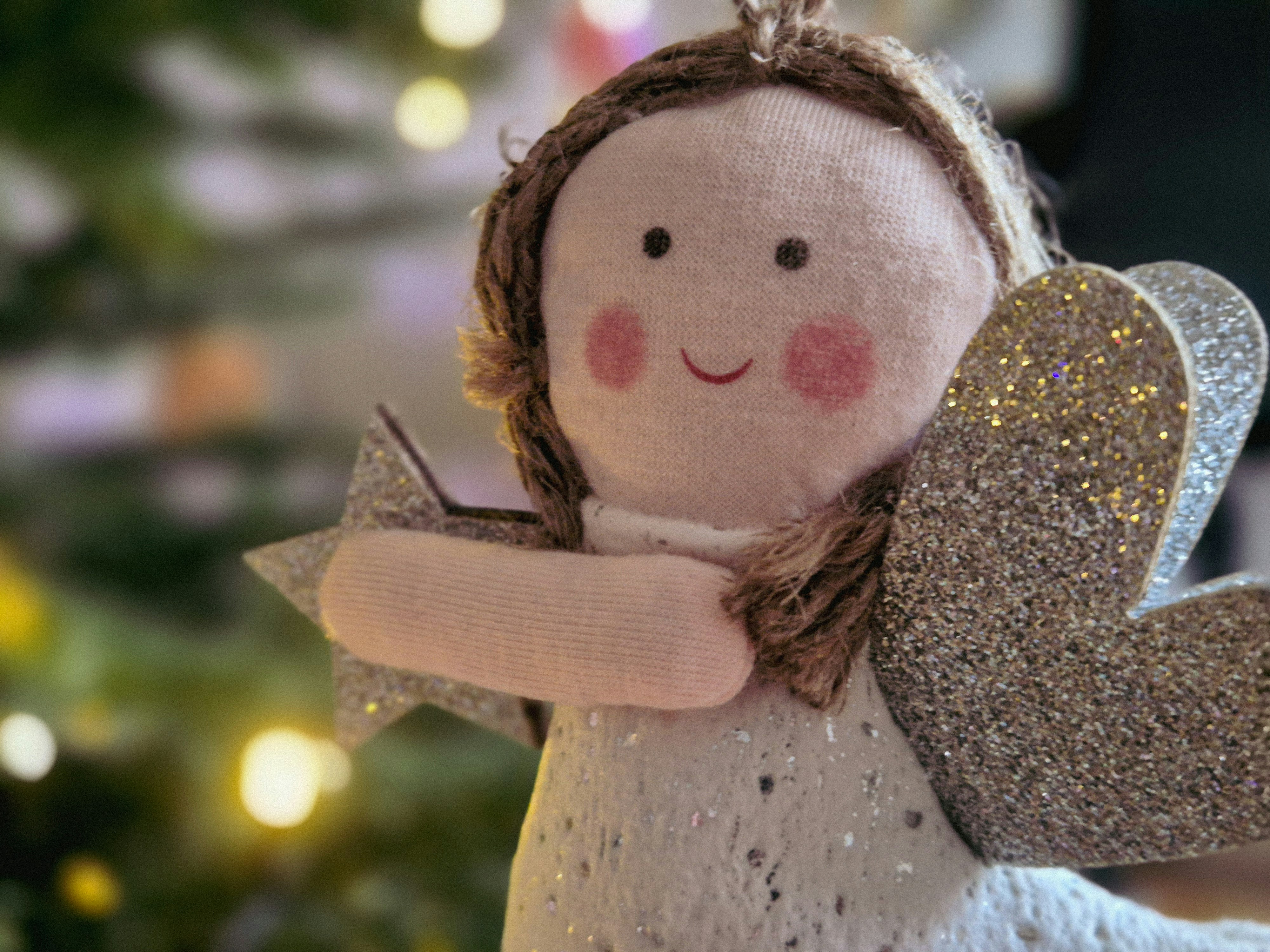 Soft fabric angel plush with glitter wings in warm bokeh glow, close-up portrait emphasizing gentle smile and rosy cheeks.