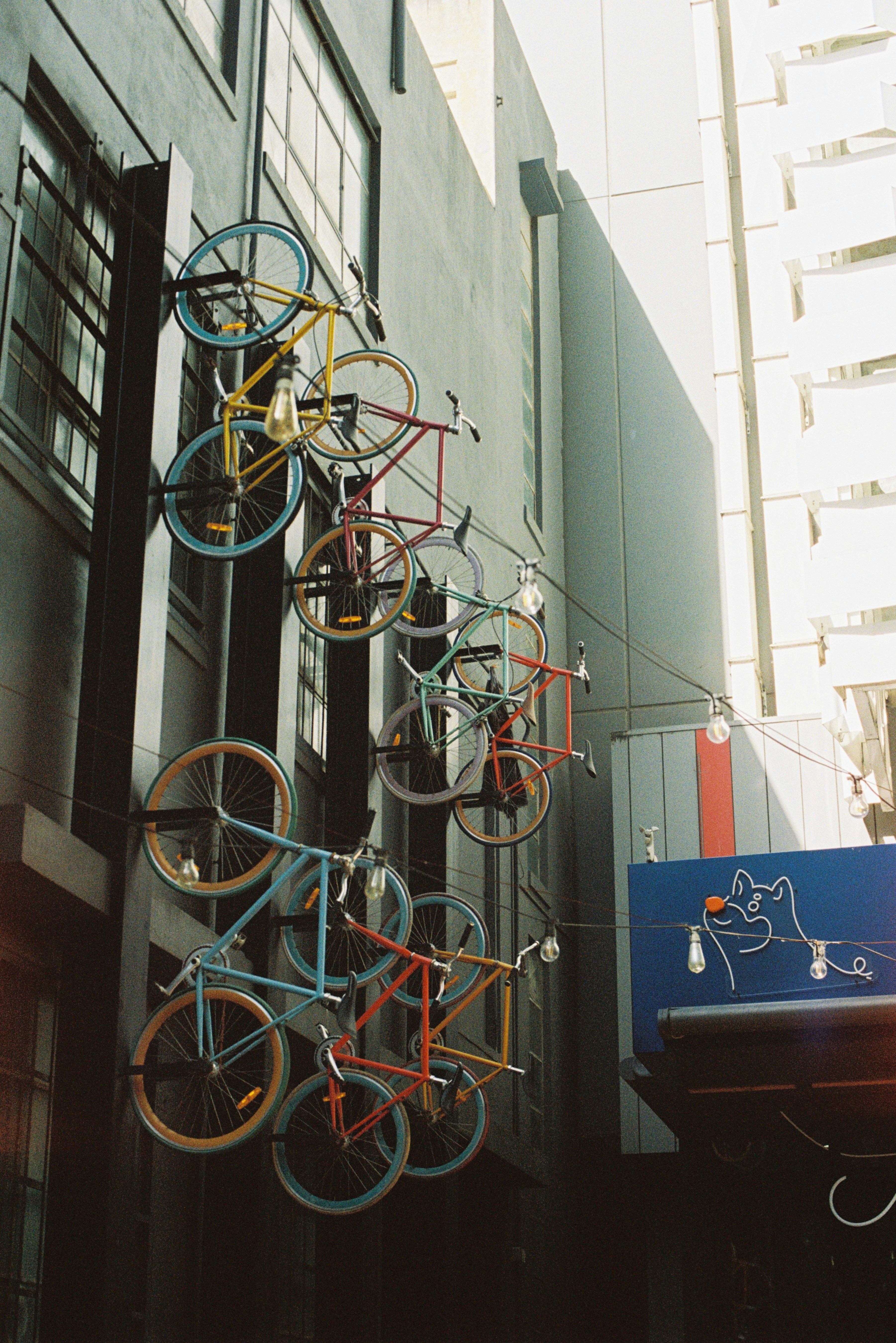 Colorful bicycles are mounted along a city wall, forming a vertical outdoor installation.