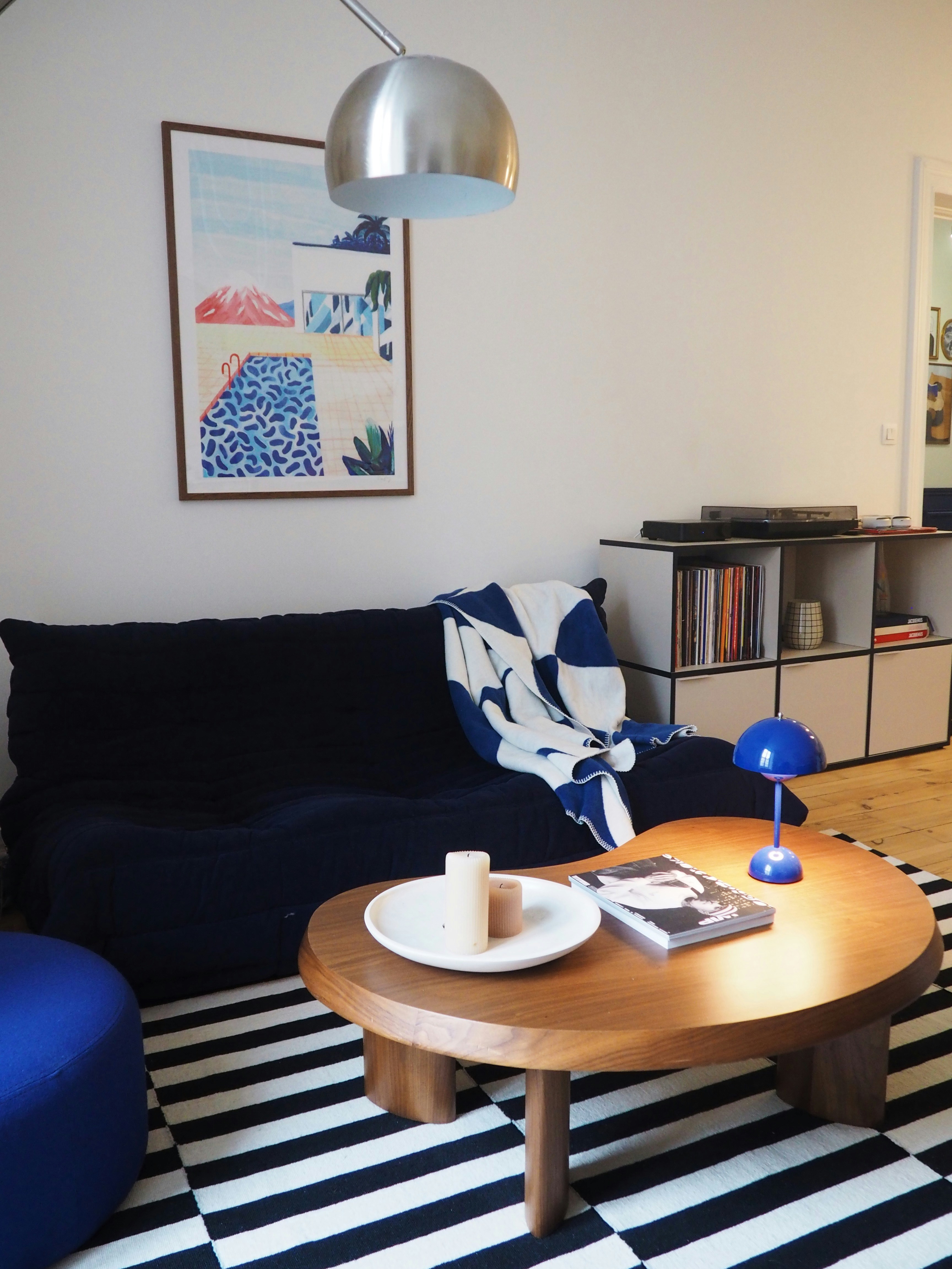 Modern living room with a black and white striped rug, wooden coffee table, deep blue sofa, and vibrant blue lamp under natural light.