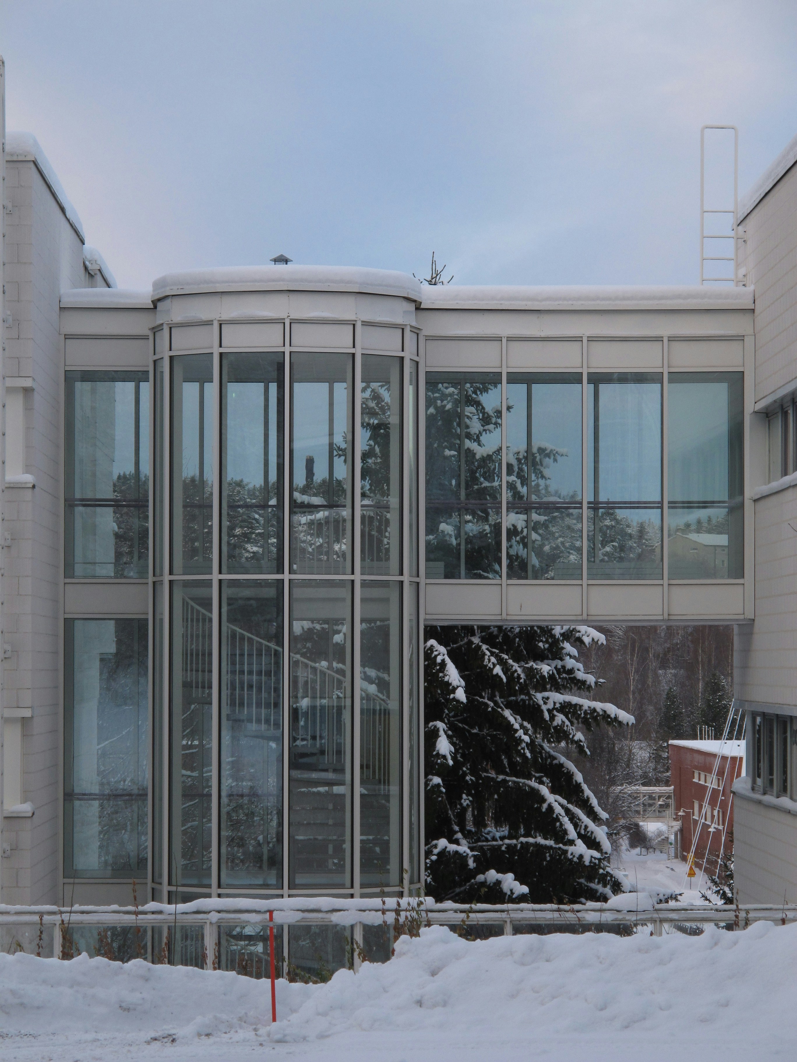 Winter daylight bathes a modern glass atrium, its curved central bay reflecting bare trees across the facade. Snow clings to the ground, framing the architectural geometry.