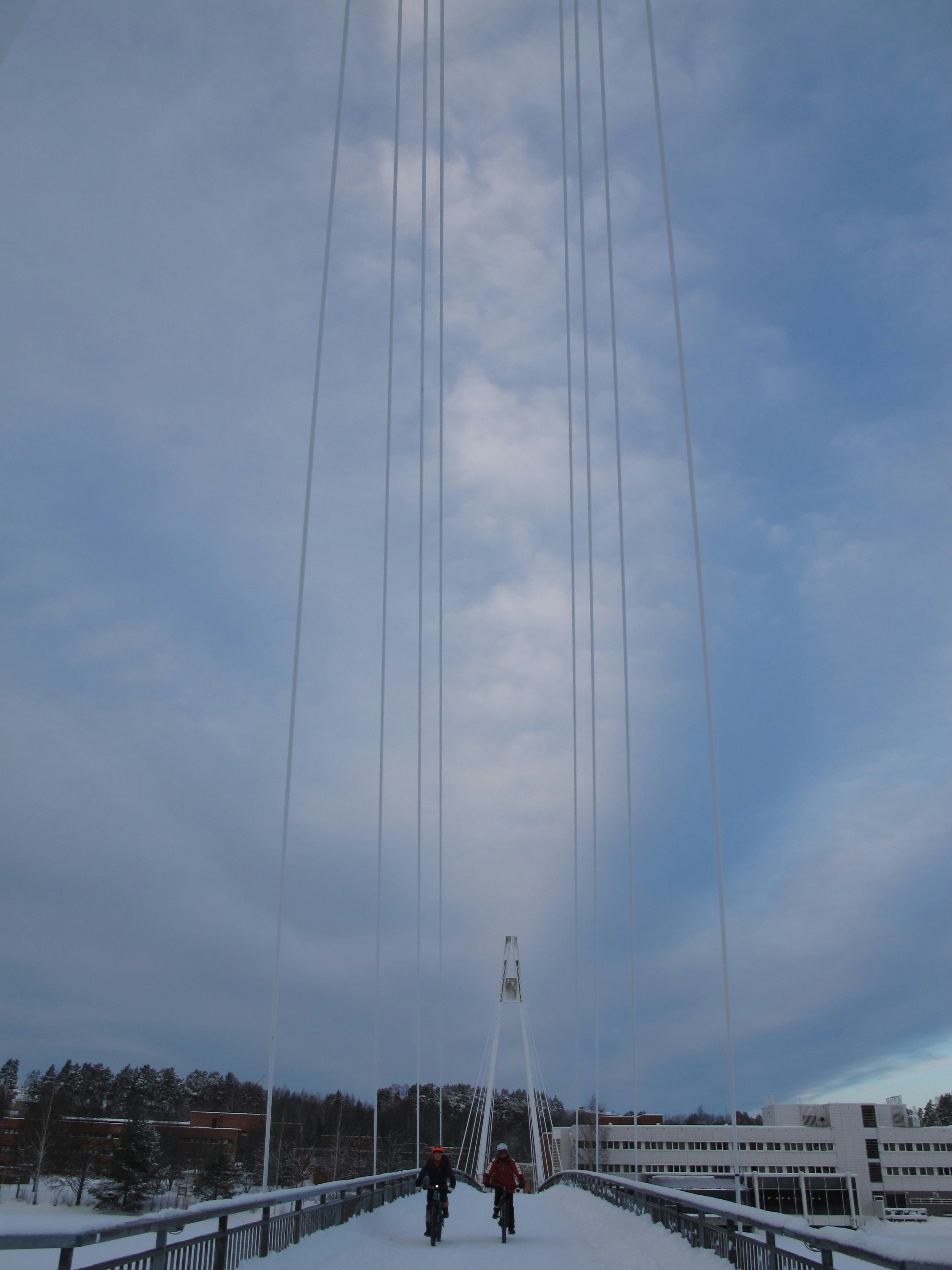 Two cyclists traverse a snow-covered pedestrian suspension bridge, framed by taut cables that rise toward a pale blue sky. Buildings line the far end, contrasting with the quiet, wintry scene.