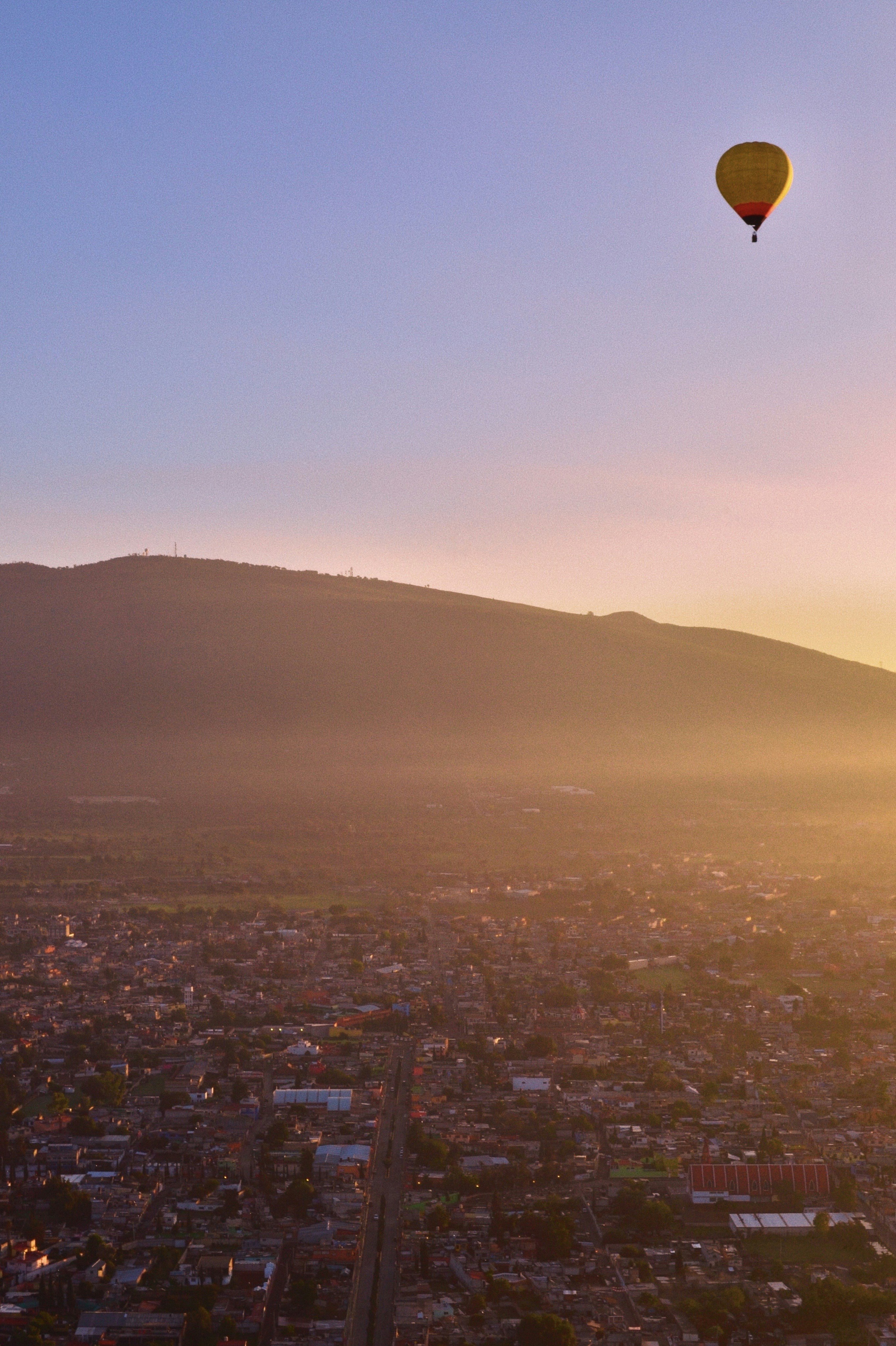 Hot air balloon drifting above a city at sunrise, with a mountain silhouette in the background.