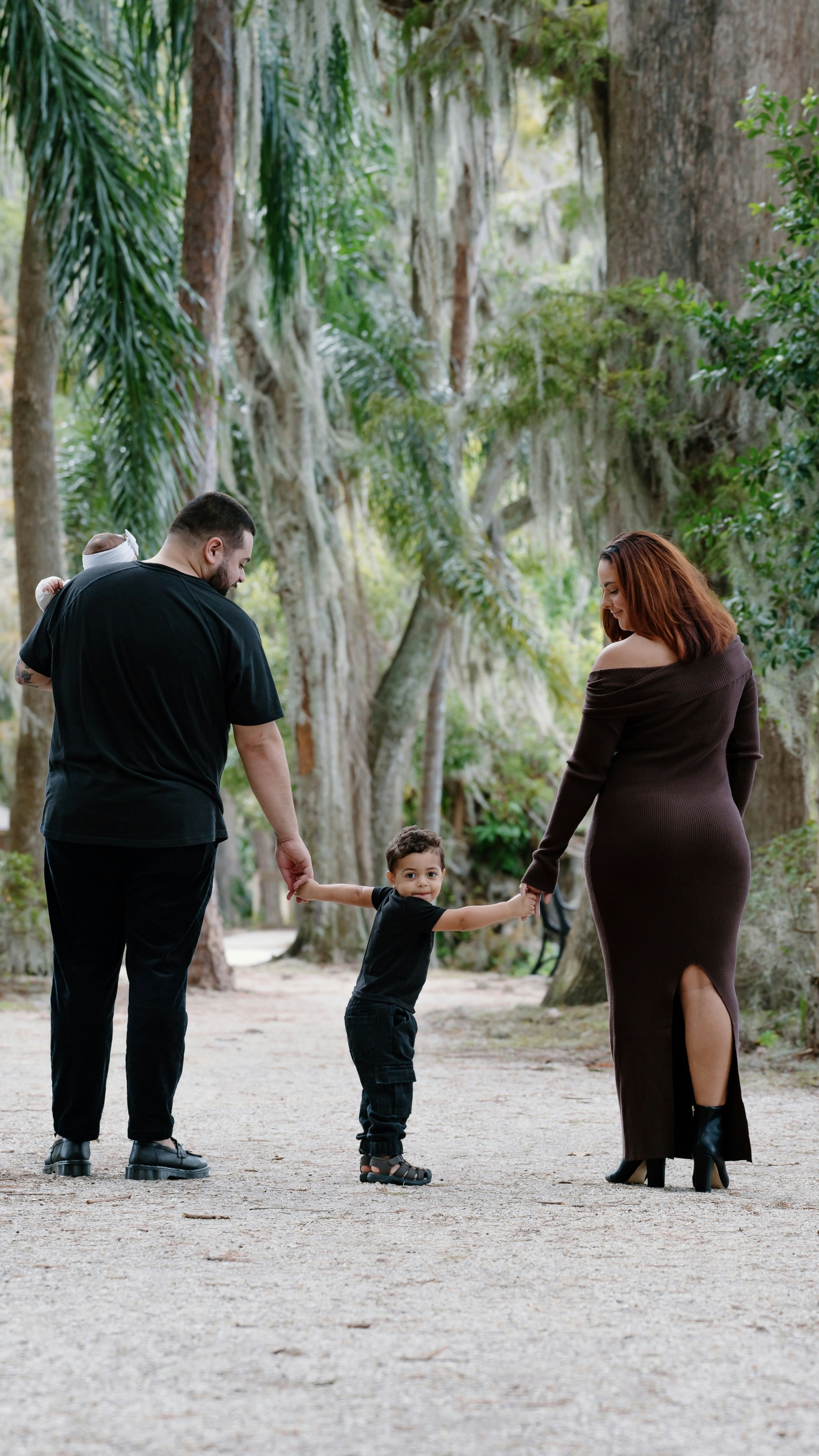 Family walking hand in hand along a forest path surrounded by tall trees and hanging moss.