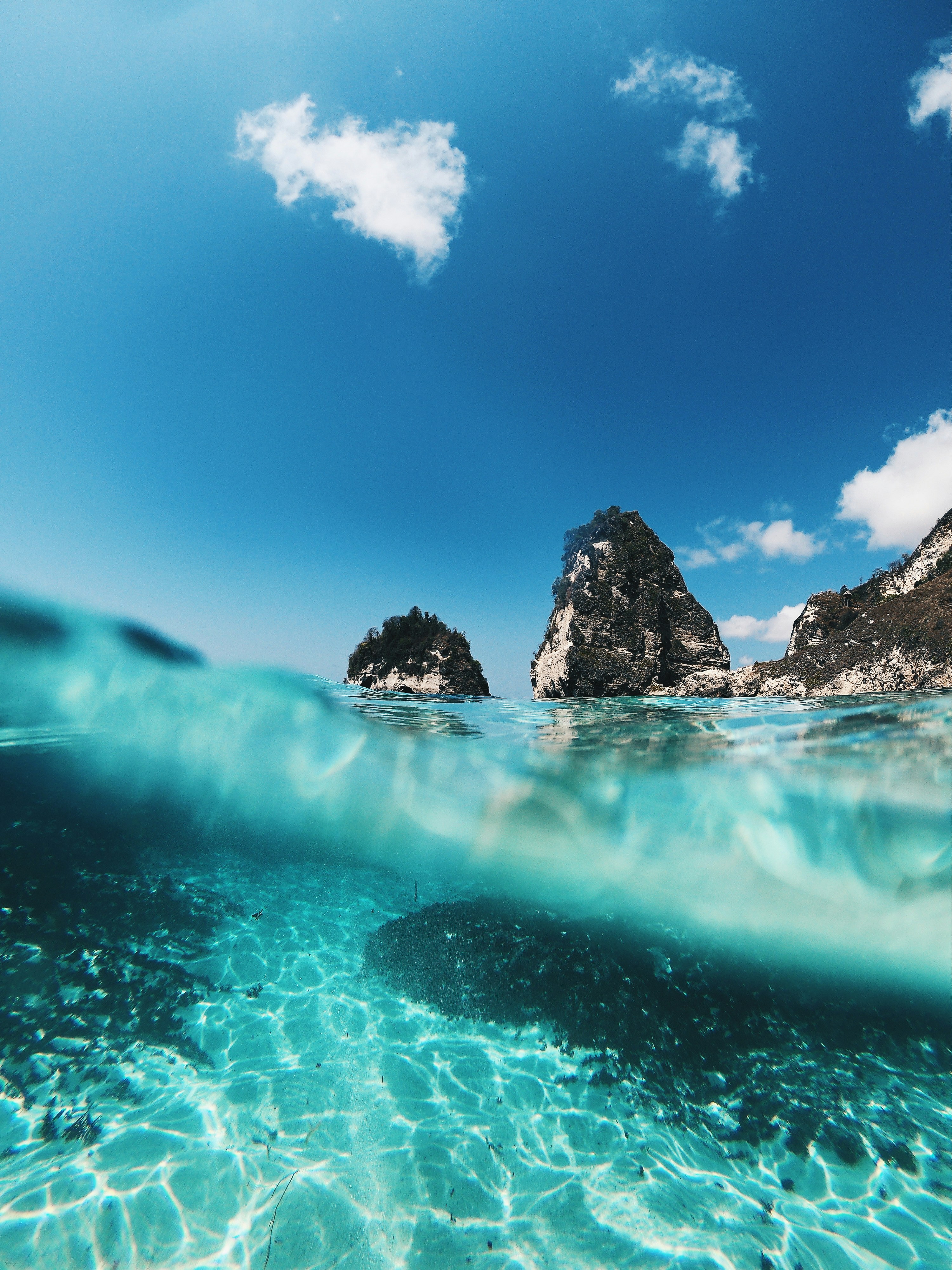 Split-level view shows turquoise water below and a bright sky above, with jagged sea stacks breaking the horizon. The image emphasizes underwater clarity meeting bold coastal rock silhouettes.