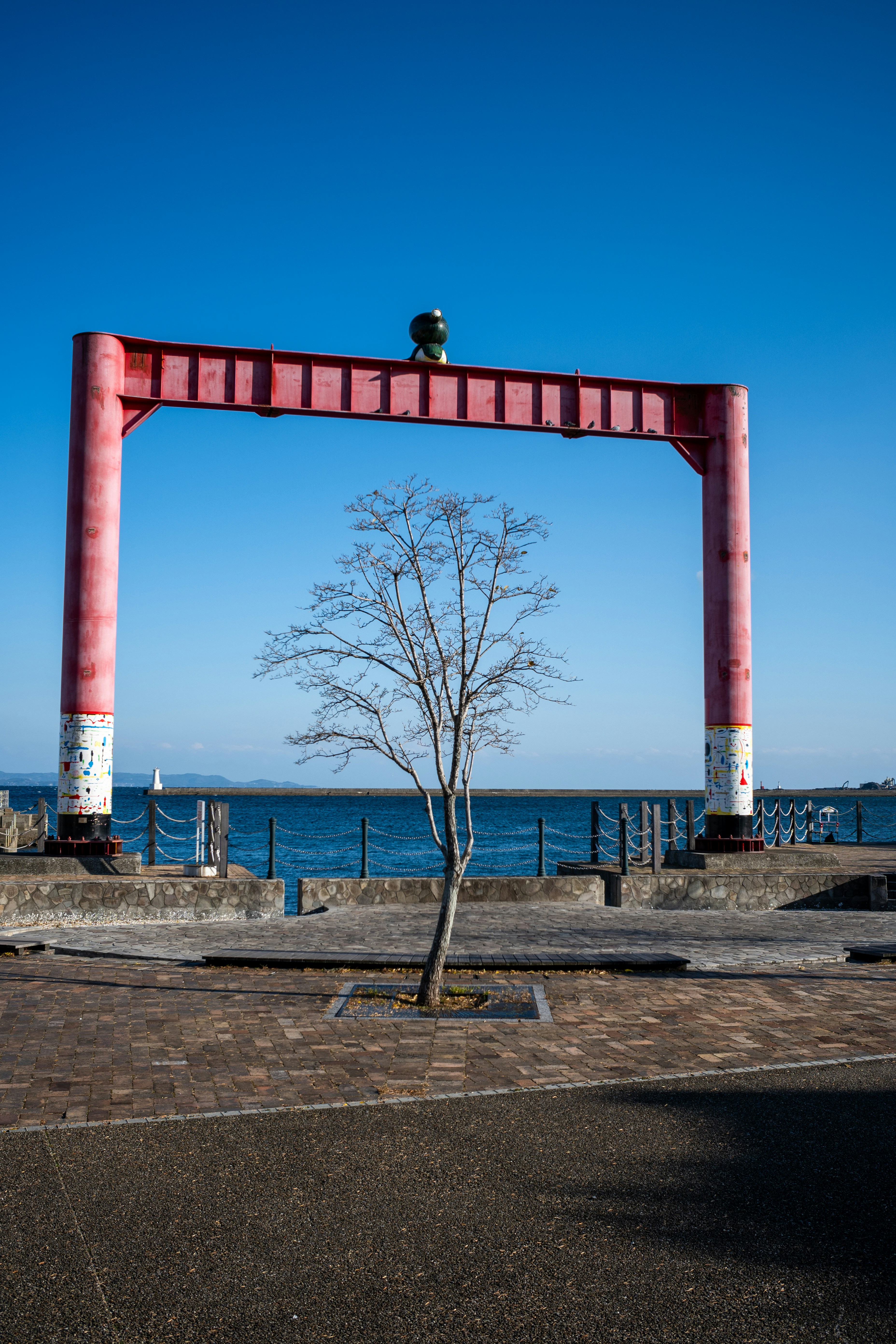 Leafless tree centered within a vivid crimson frame by the harbor, with a calm sea and bright blue sky behind.