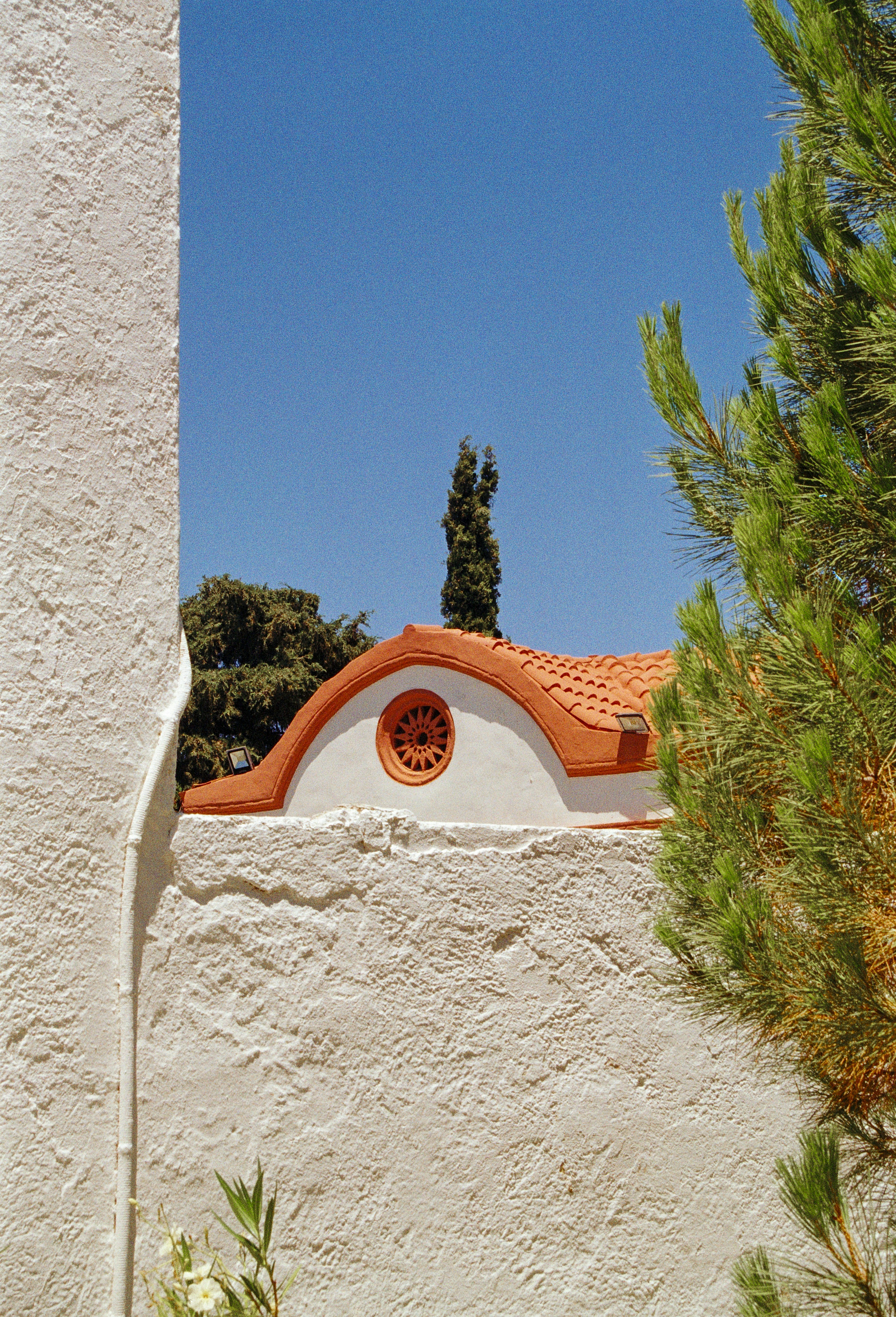 Mediterranean-style wall with a circular vent beneath an orange-tiled arch, set against blue sky and green foliage.