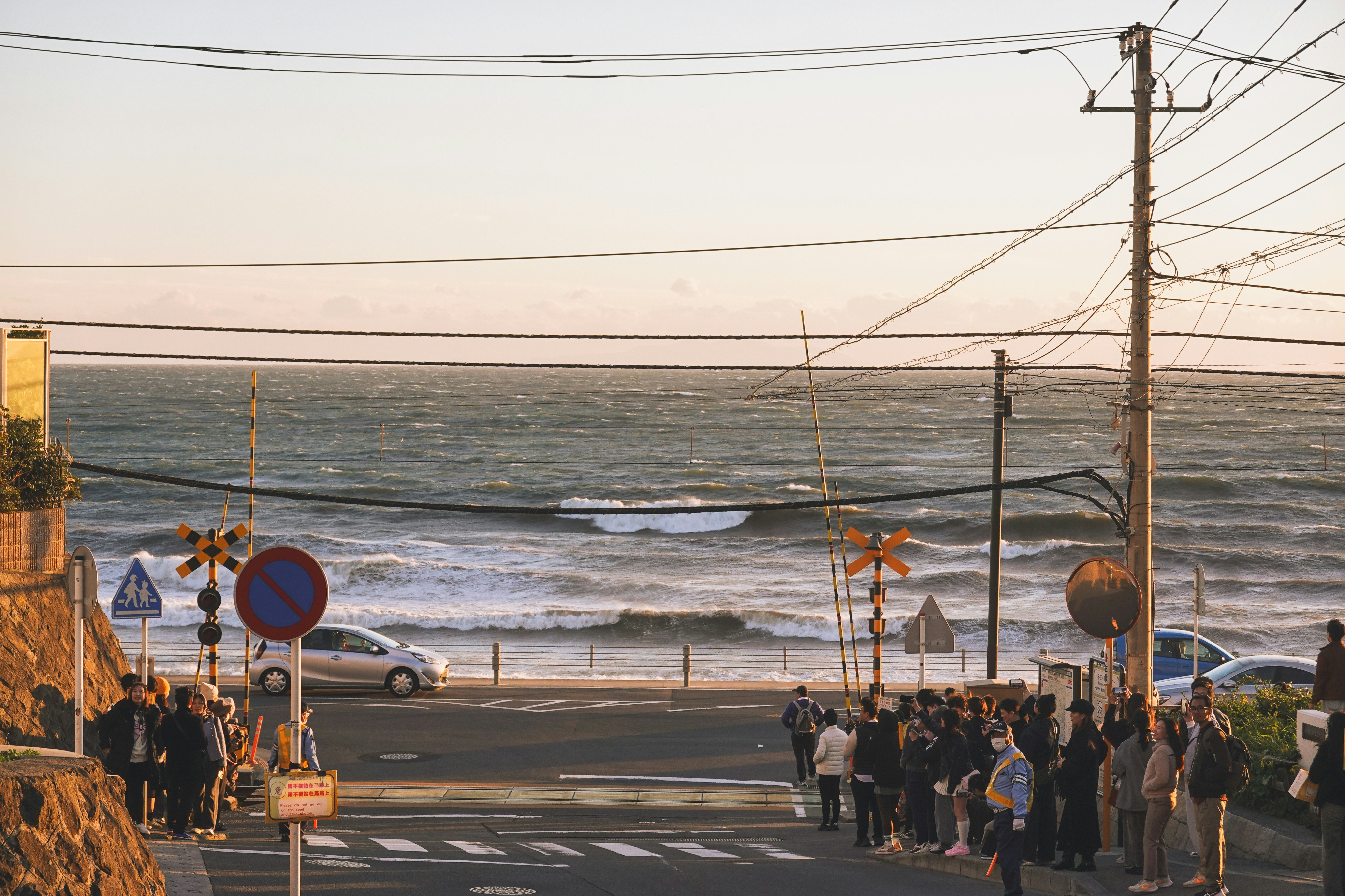 People gather near a coastal road with ocean waves breaking under a warm sunset sky.