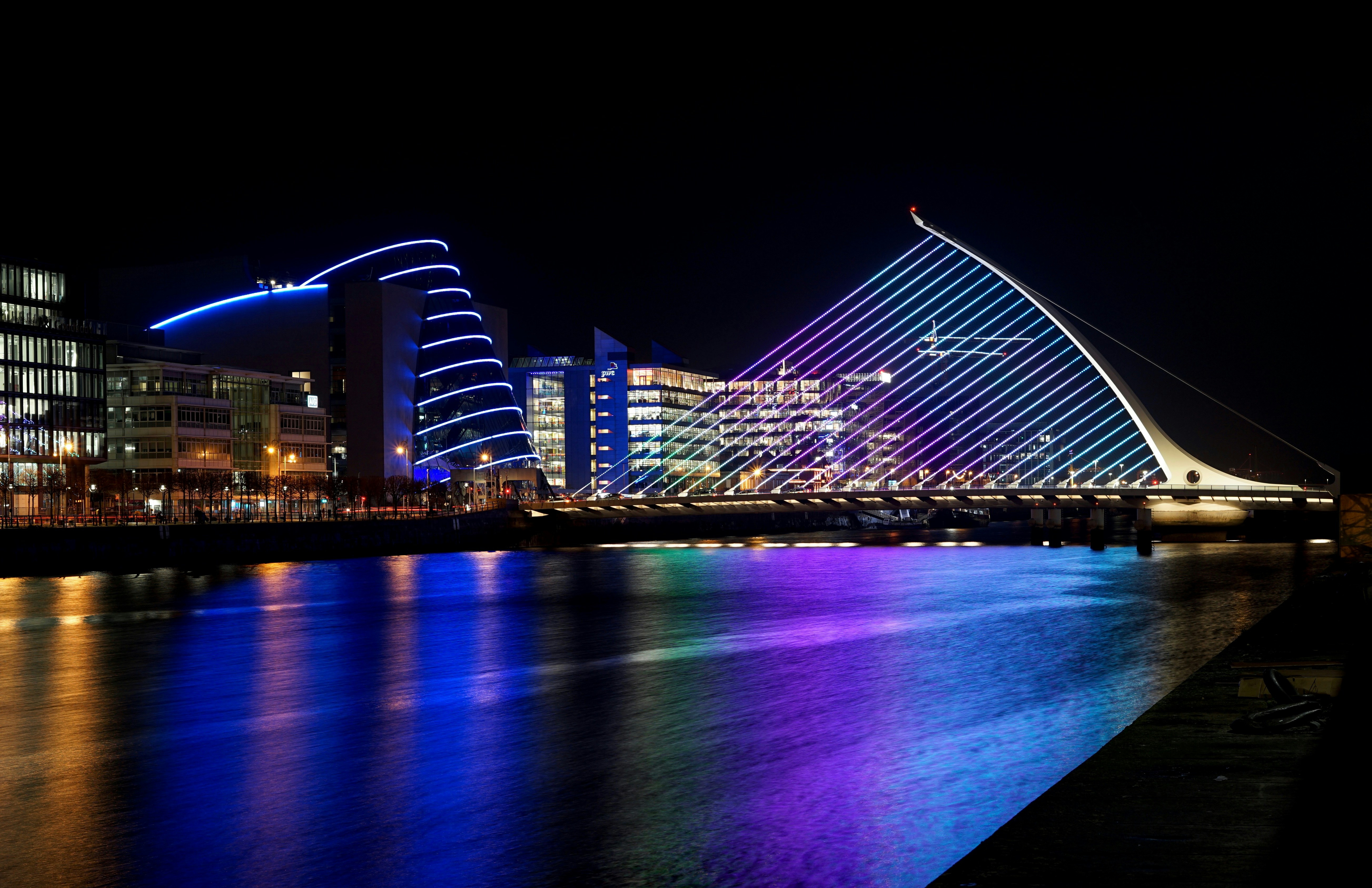 Vibrant city lights reflecting in the tranquil river at night, featuring a uniquely designed illuminated bridge.