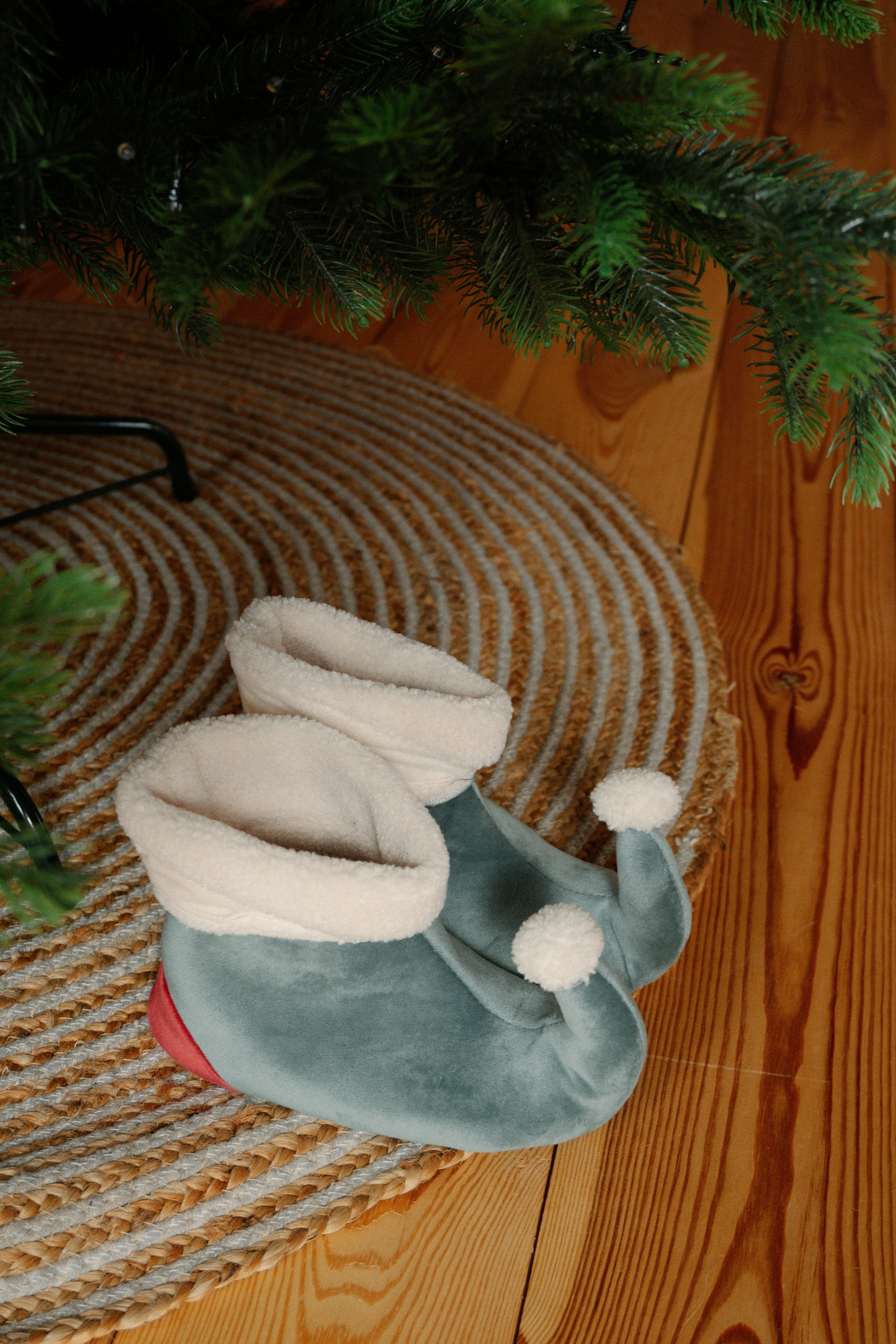 Pastel blue velour slippers with fluffy white trim rest on a circular rope rug over a warm wooden floor, with evergreen branches overhead.