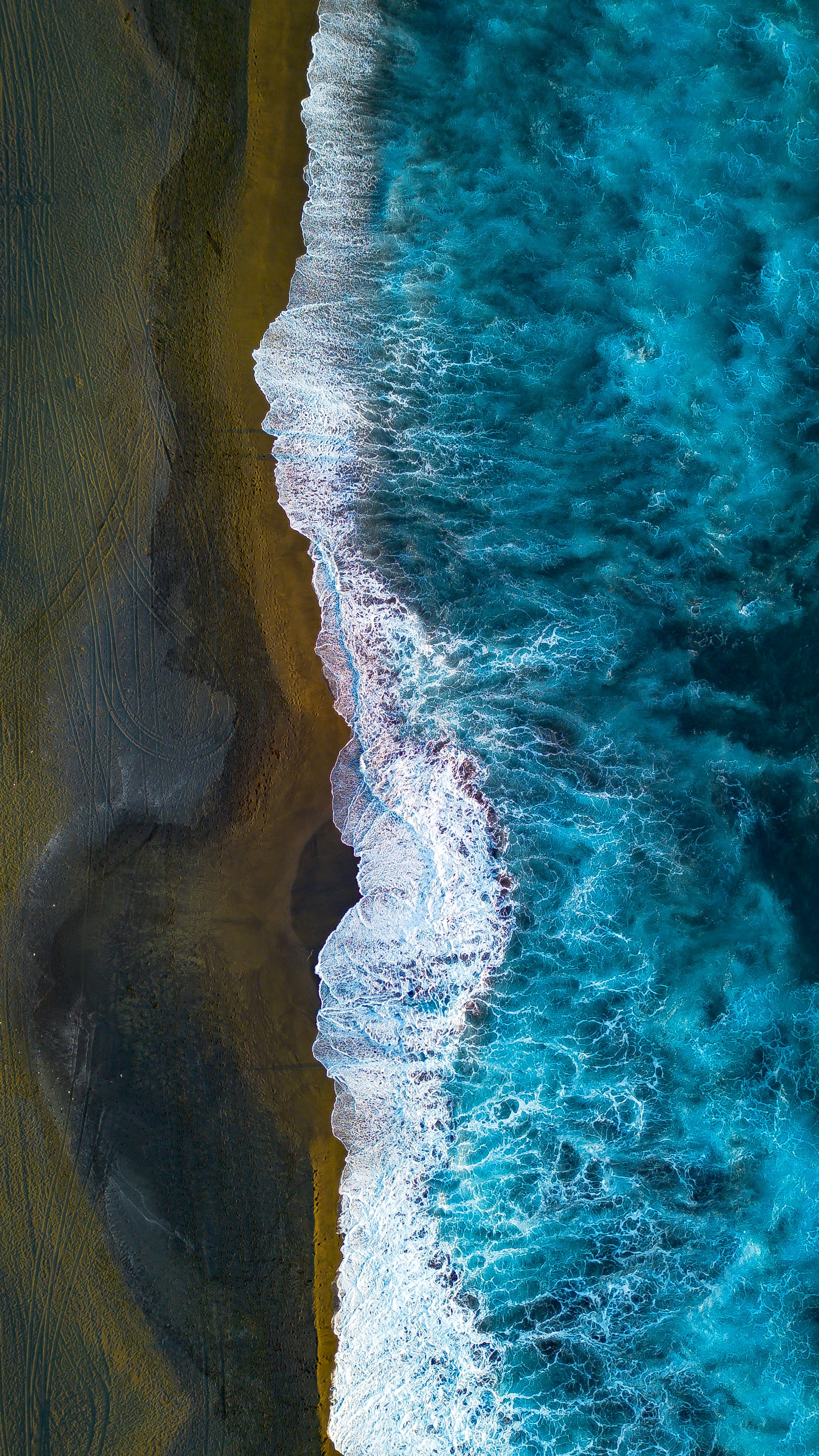 Vibrant turquoise waves crashing onto a sandy beach, creating a dynamic contrast with the darker coastal landscape. 