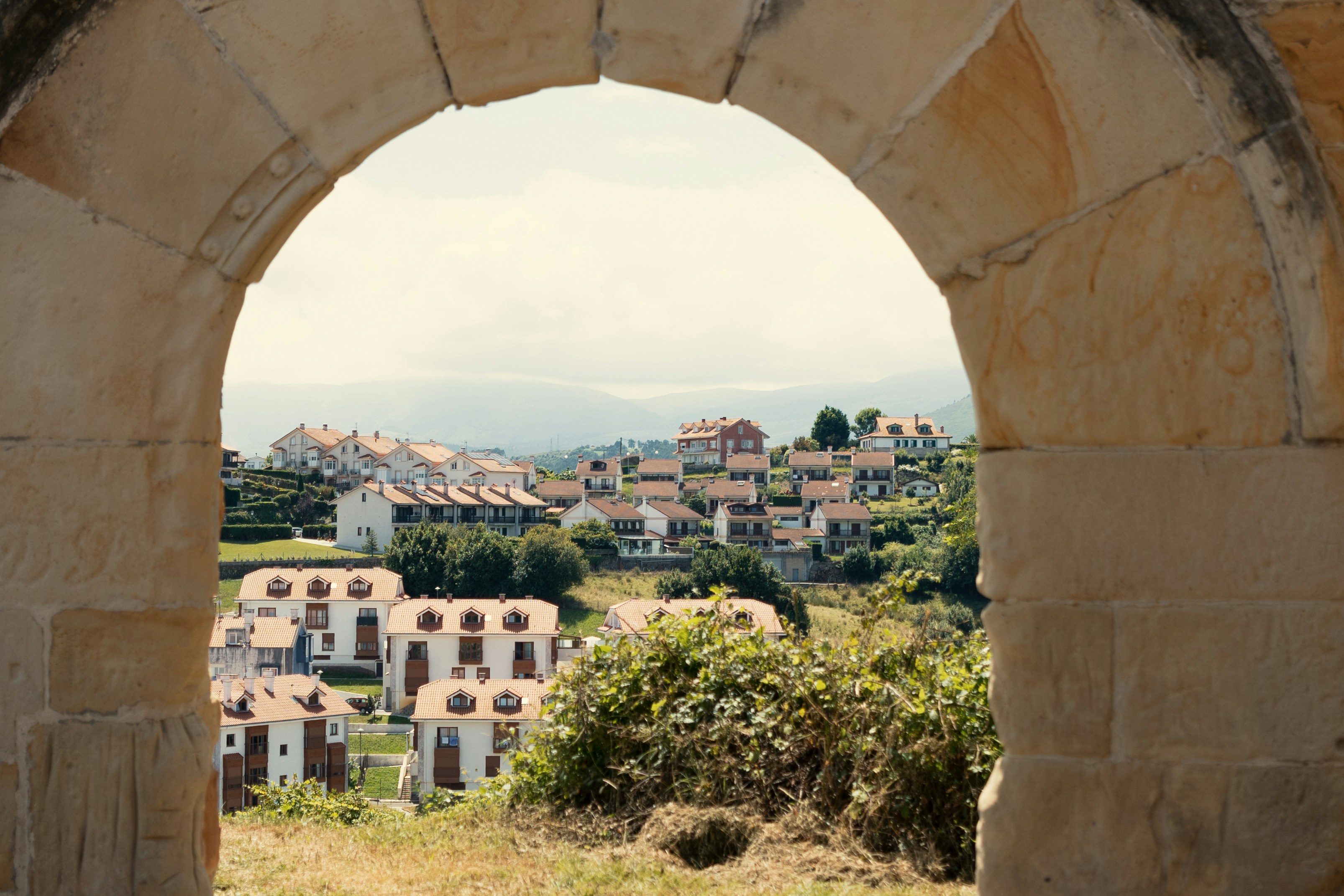 Stone arch frames a view of a village with tiled rooftops and green hills under a cloudy sky.