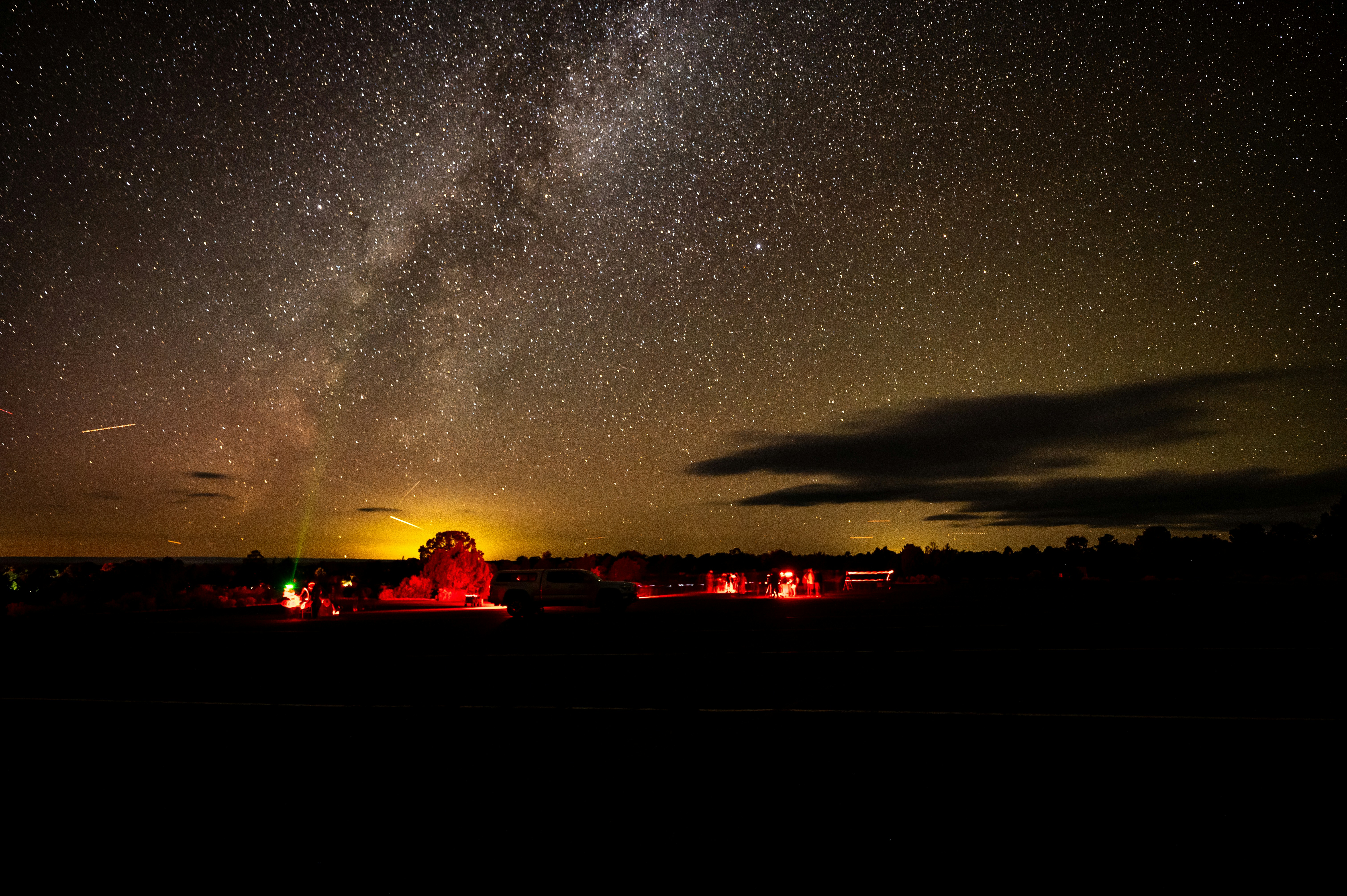 Milky Way arches across a star-filled night sky above a softly illuminated campsite, with warm red tones accentuating the silhouettes of trees and tents.