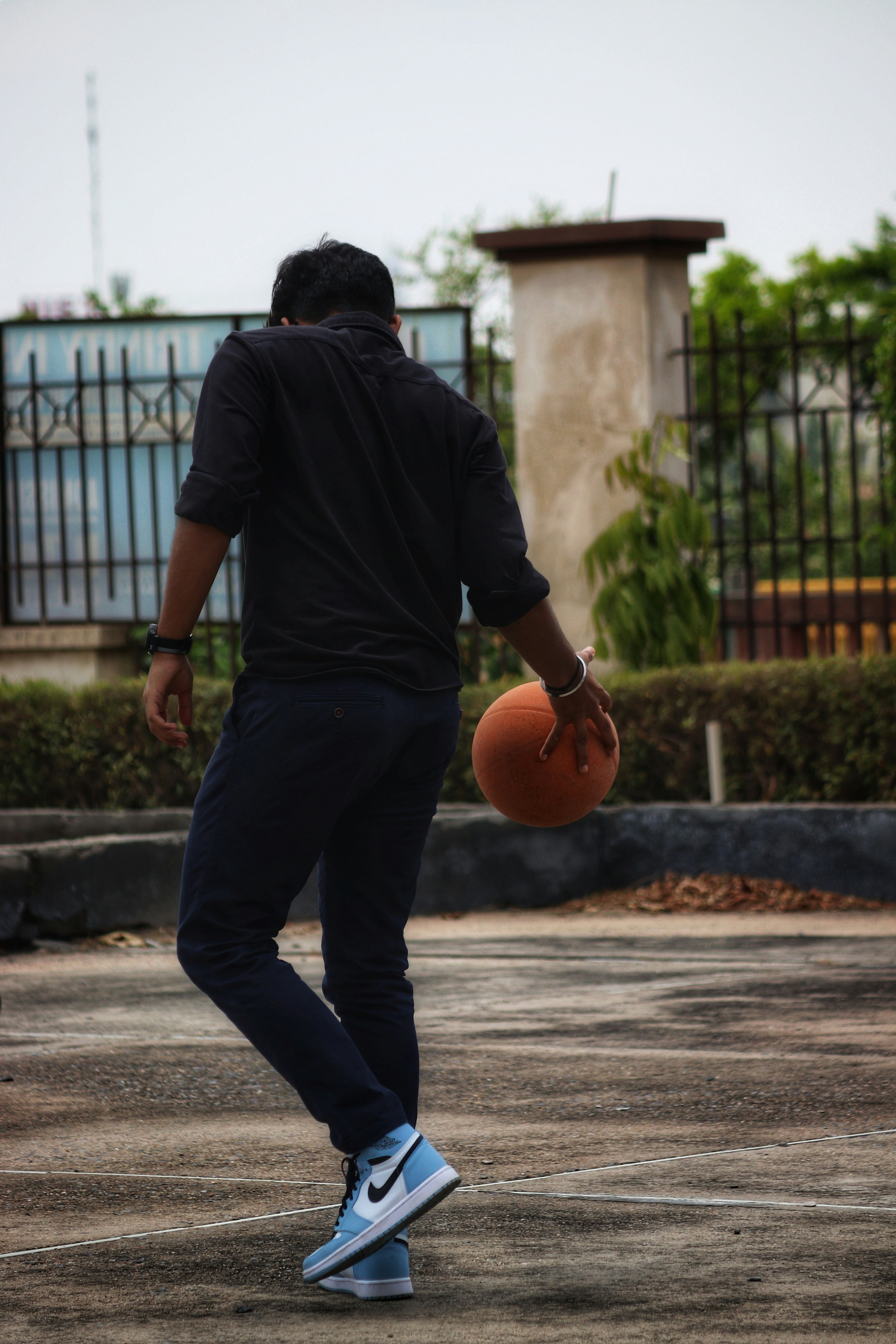 A young man in a black shirt and blue sneakers stands on a basketball court, holding a ball and gazing thoughtfully into the distance.