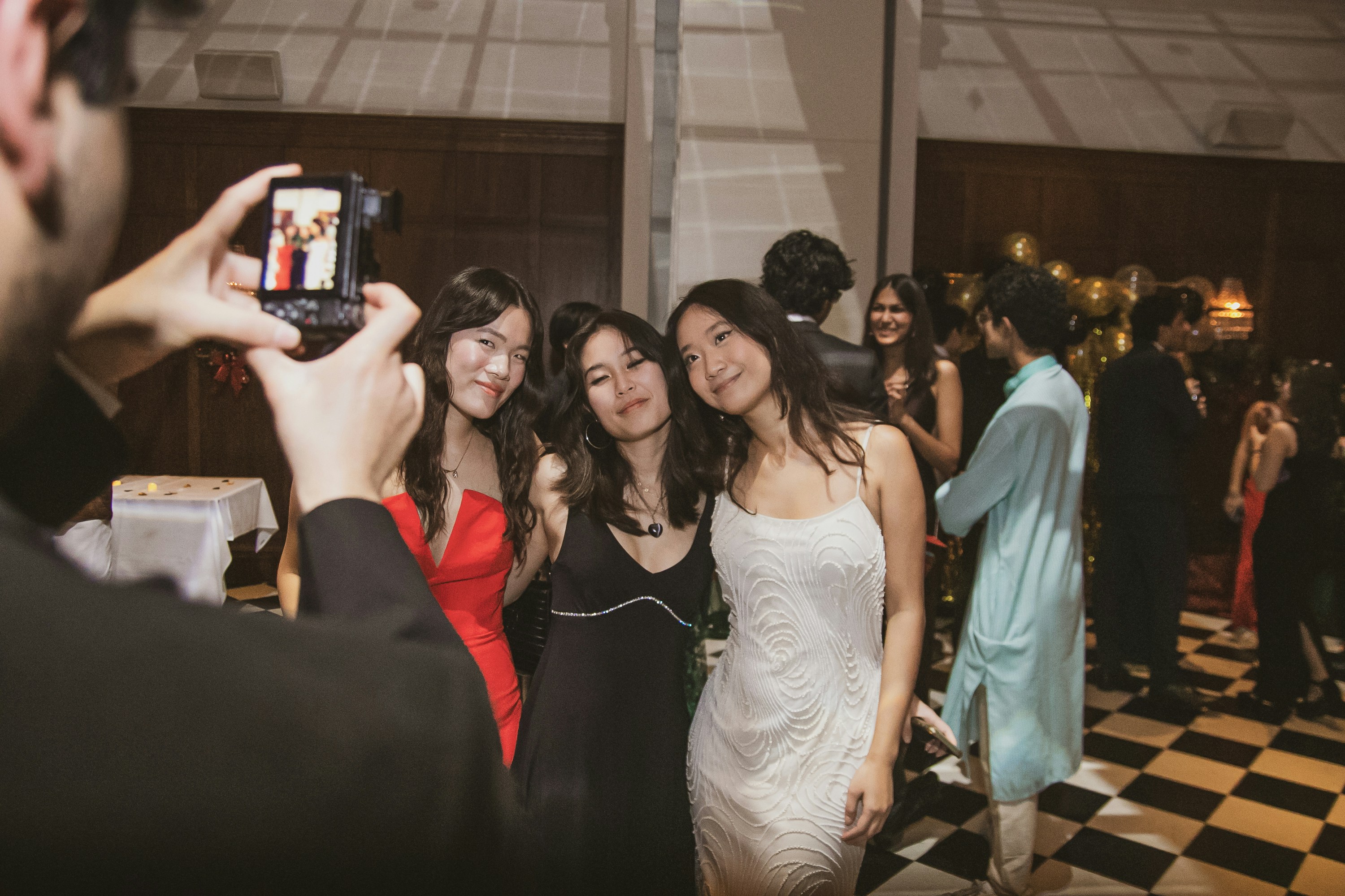 Three elegantly dressed individuals posing for a photo at a formal event, with a lively crowd in the background.