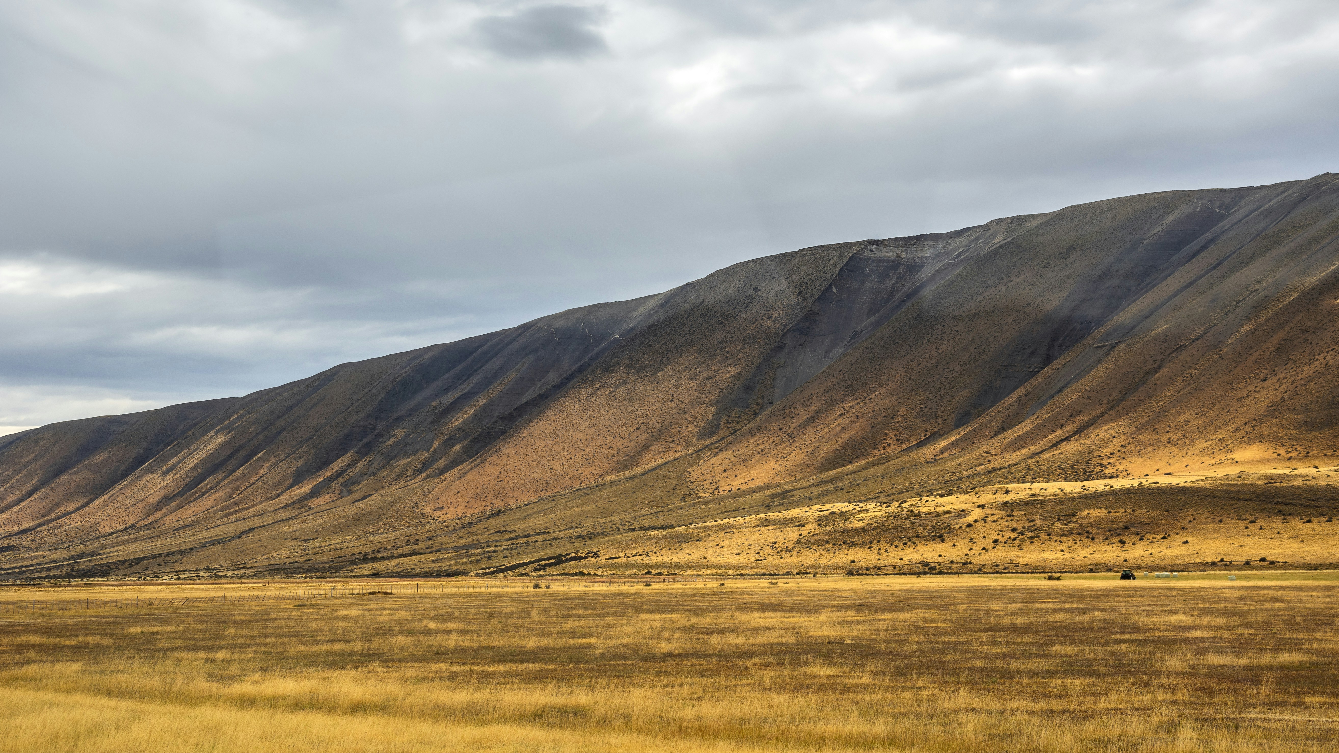 Expansive grassland meets the shadowed slopes of a rugged mountain range under a moody sky.