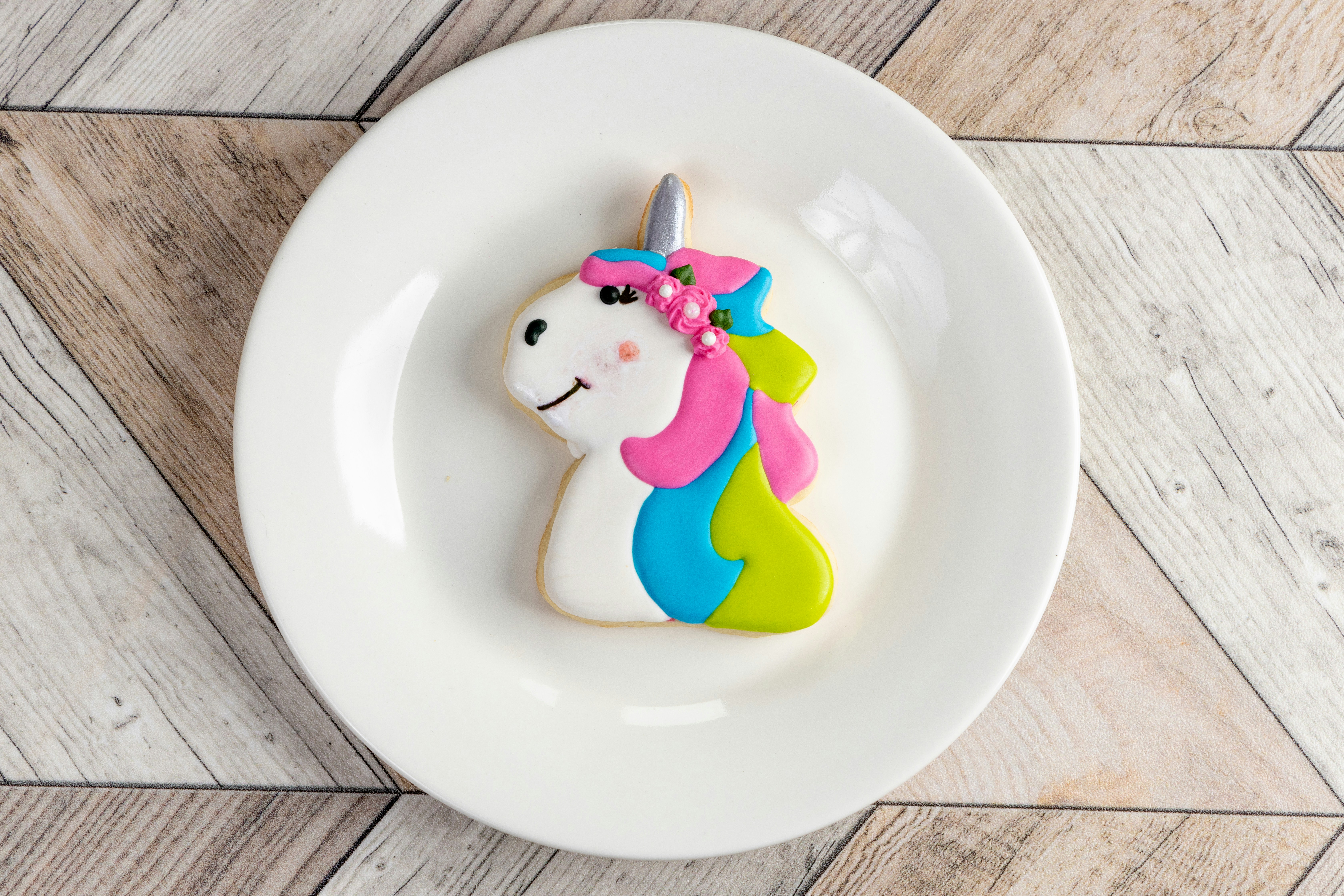 Colorful unicorn-shaped cookie on a white plate against a wooden backdrop.