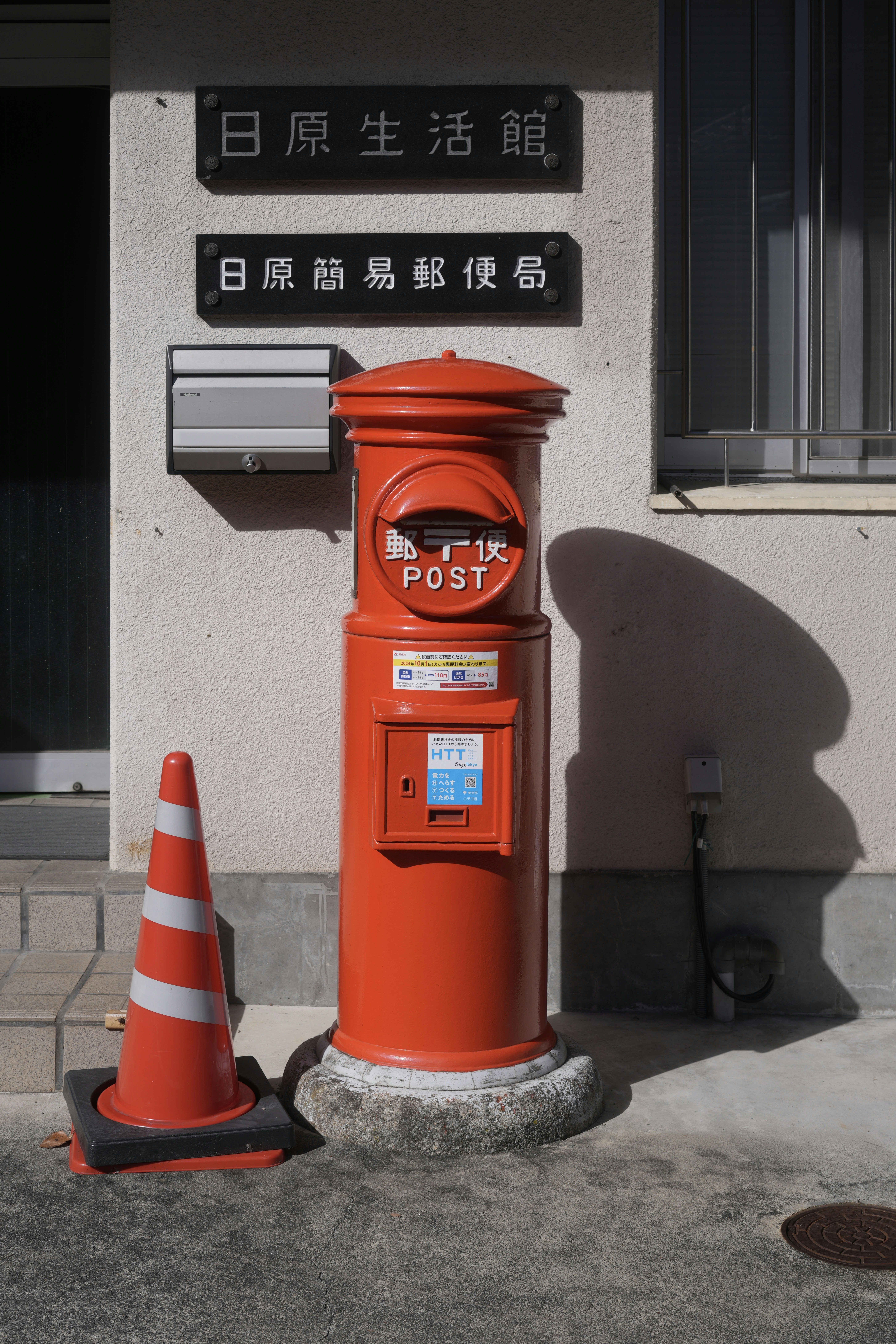Bright red Japanese mailbox standing prominently beside a traffic cone and building facade, showcasing urban postal heritage.
