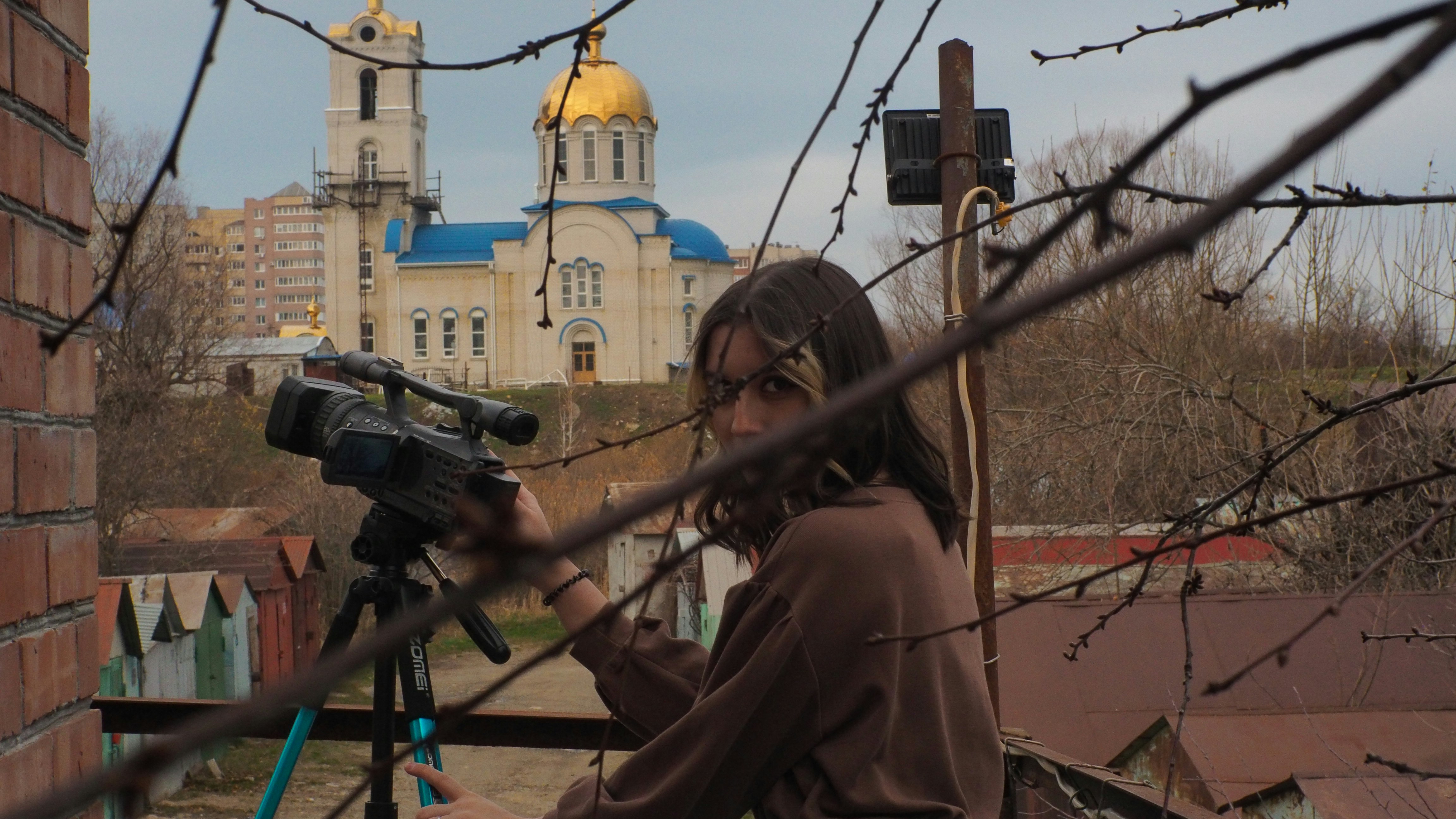 A street videographer peers through foreground wires while filming a cityscape dominated by a blue-domed church.