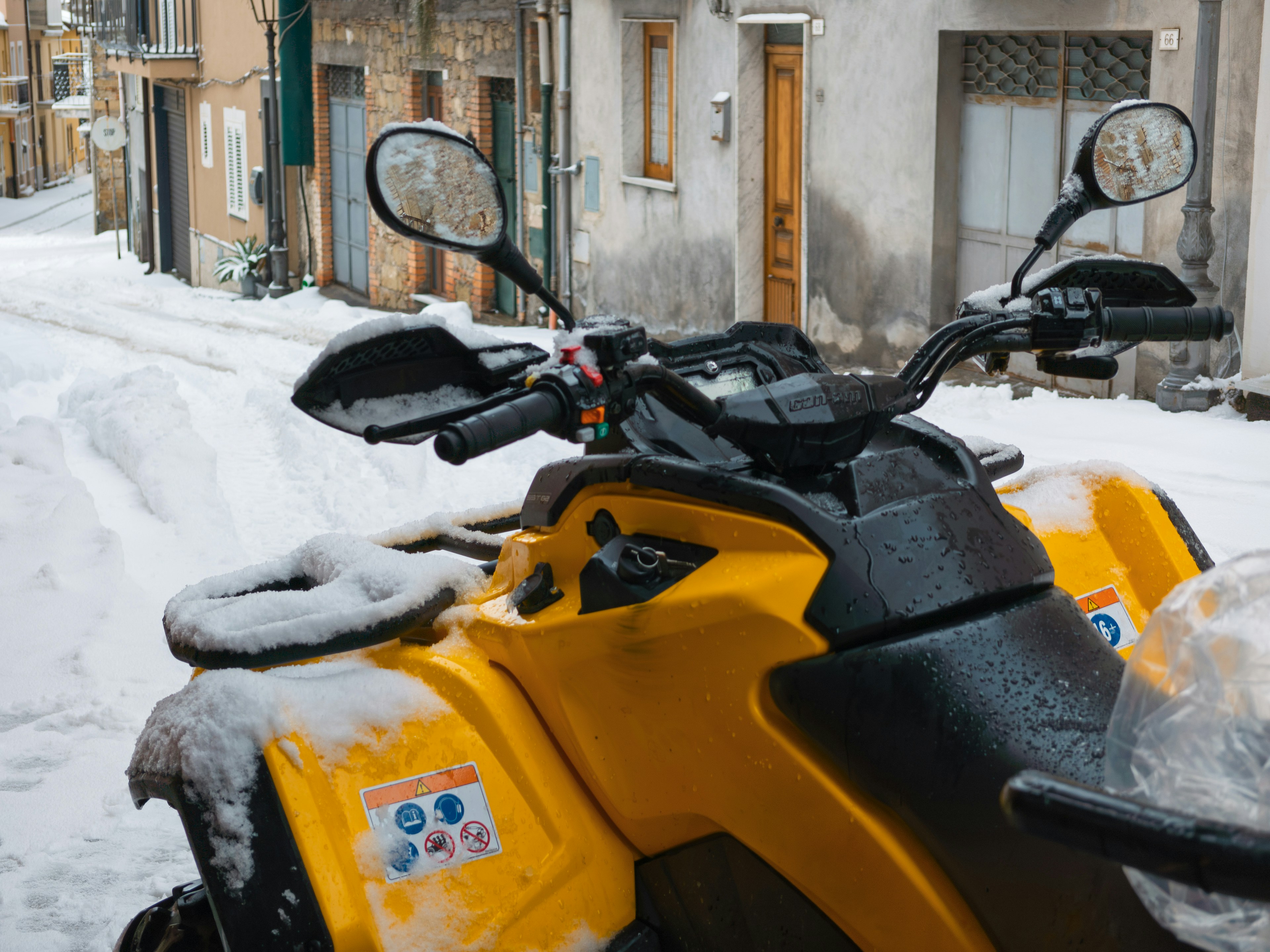 Yellow scooter sits on a snow-covered street, with damp snow on the seat and handlebars as weathered buildings line the lane.
