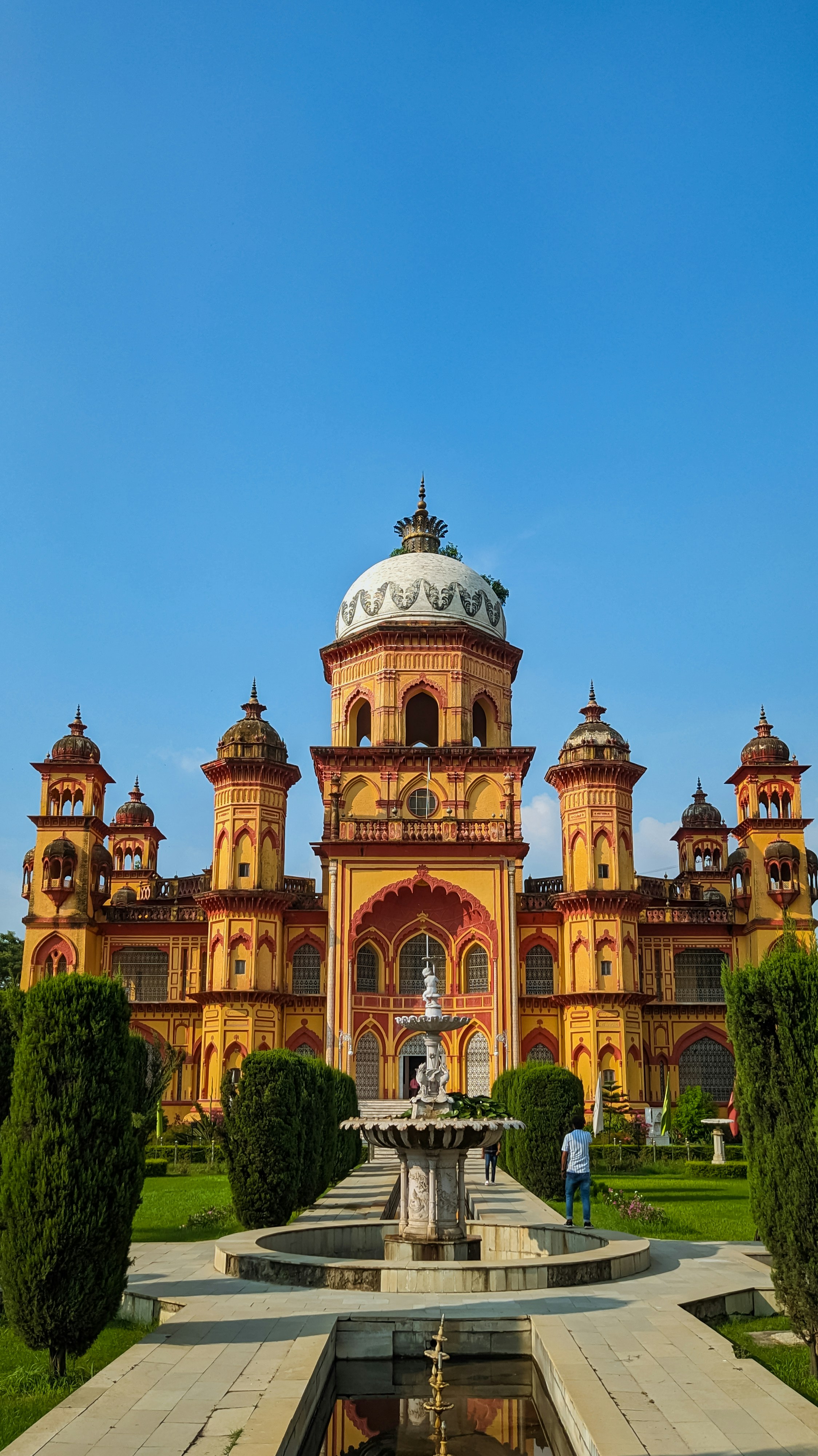 Colorful palace complex with a central fountain and manicured grounds, showcasing intricate architecture and blue sky. This is a photograph of a historic building with a formal courtyard.