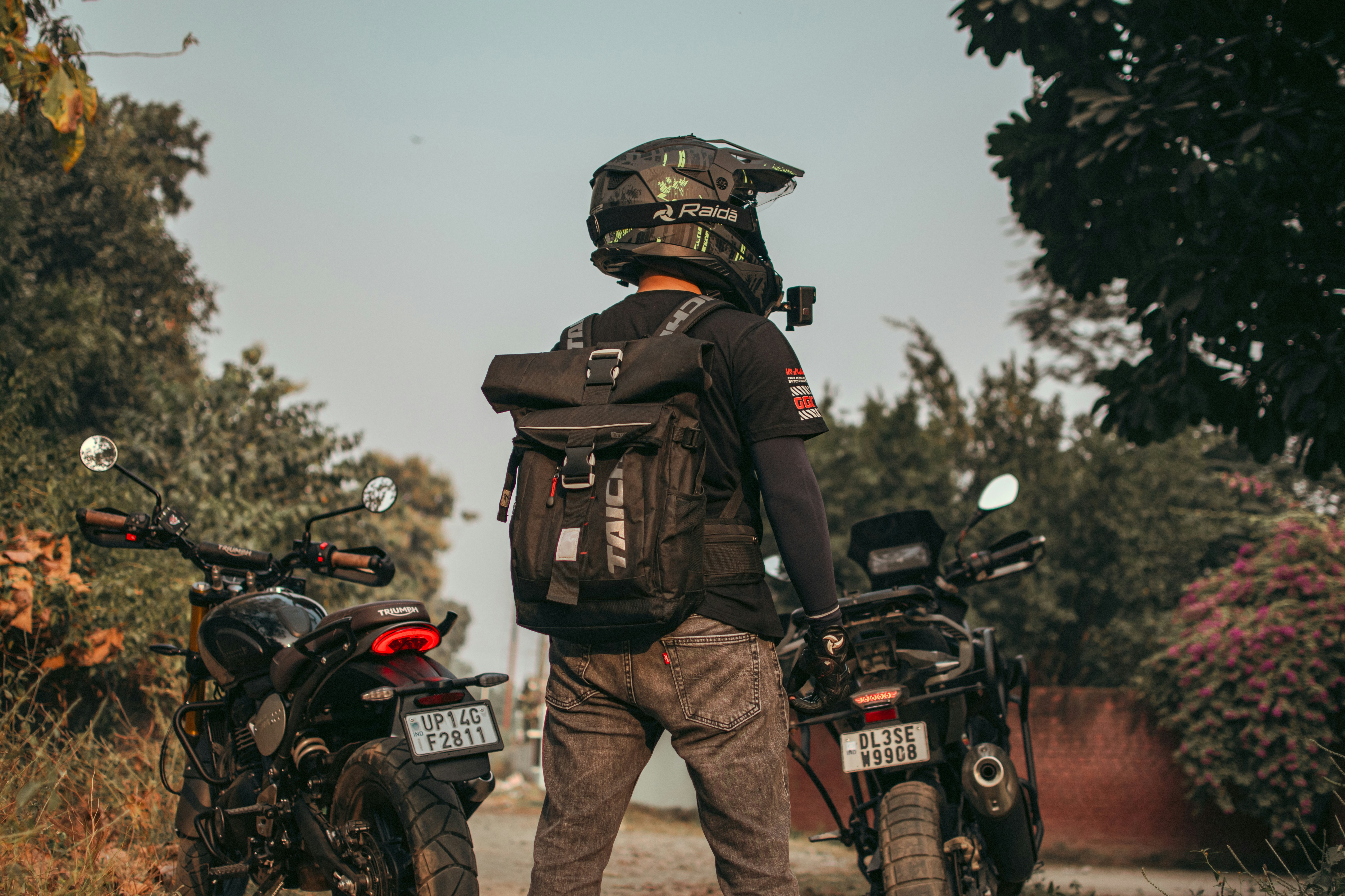 A motorcyclist stands at a crossroads, flanked by two bikes, contemplating the adventure ahead. The scene is set against a backdrop of lush greenery and vibrant flowers.