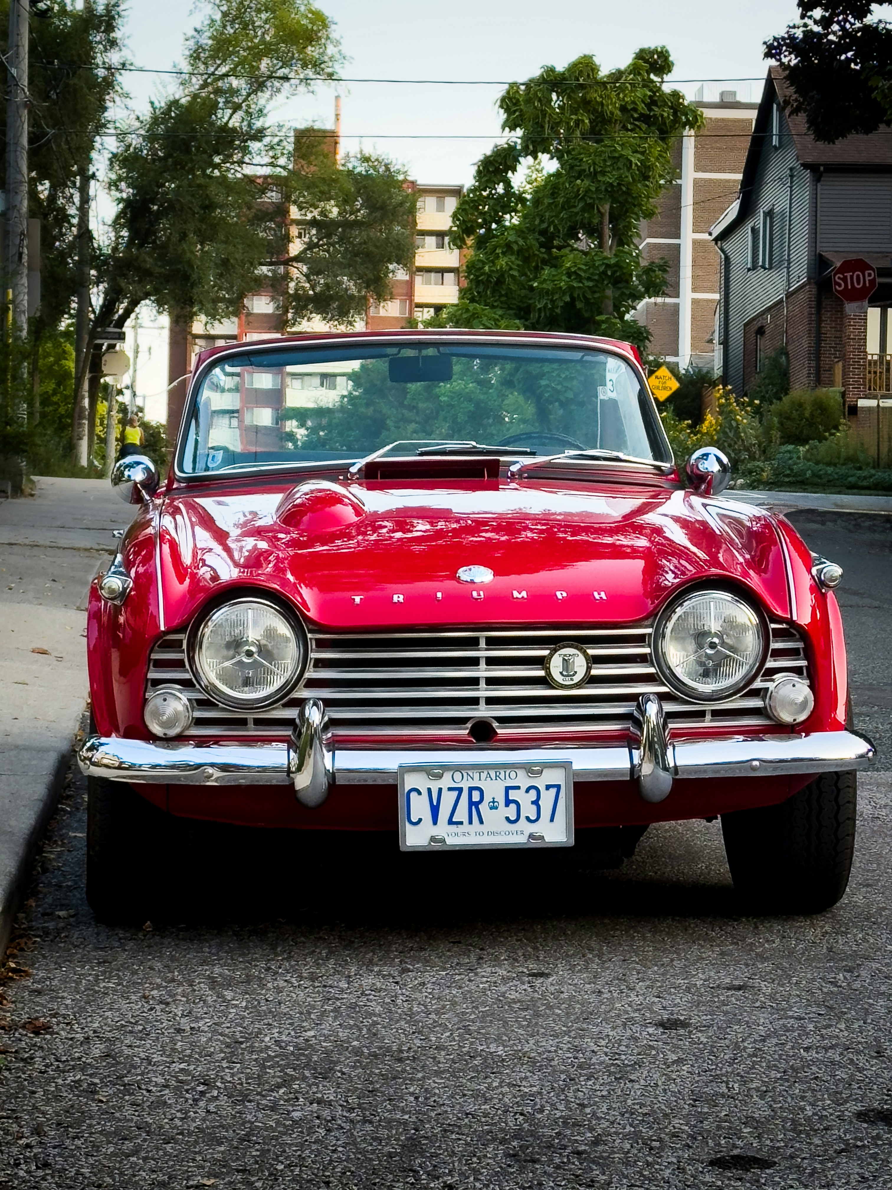 Bright red Triumph sports car parked on a city street, showcasing its vintage design and distinctive features. The backdrop includes urban elements and greenery.