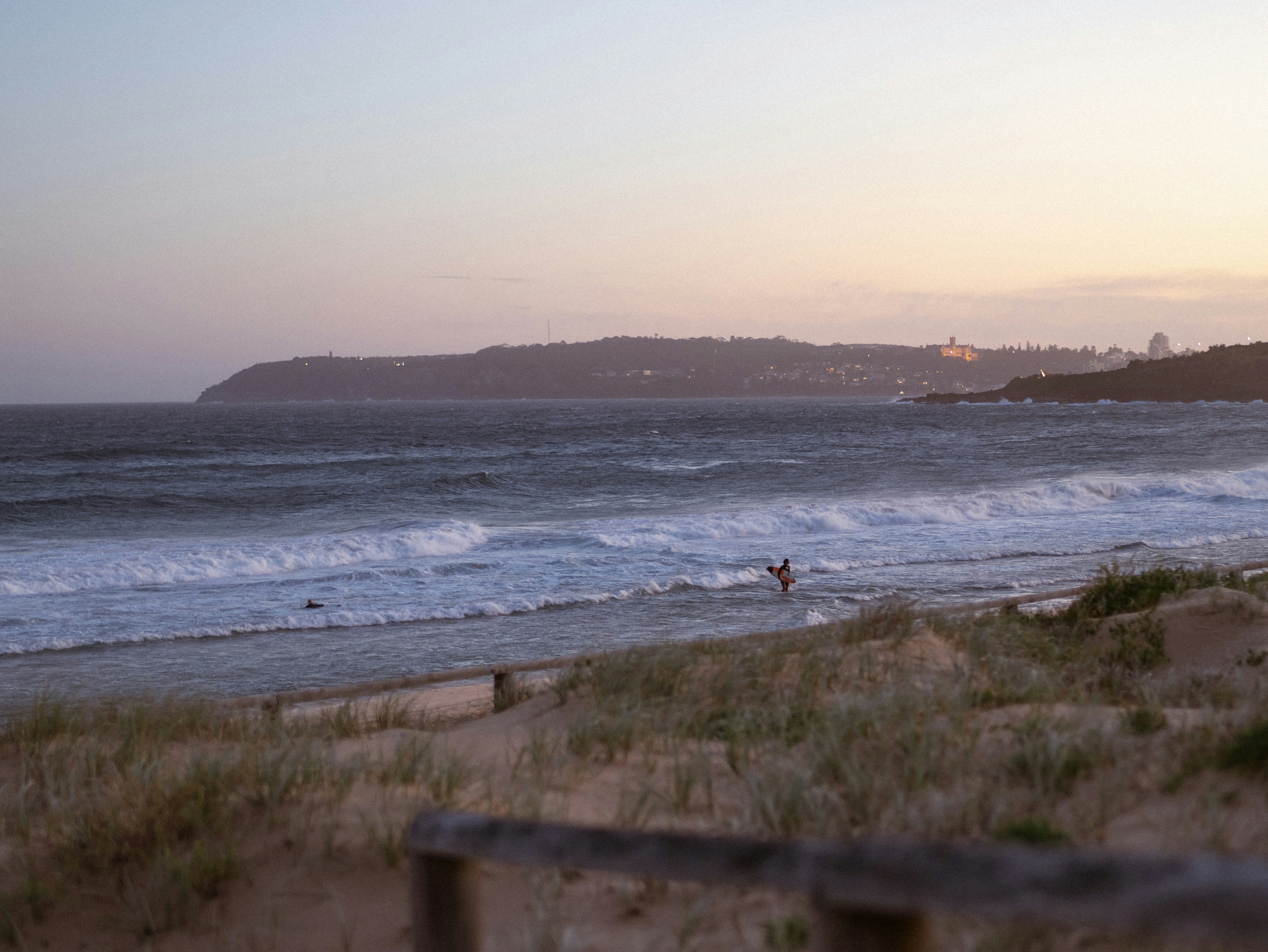 Person wading into gentle surf at dusk with sandy dunes and pastel sky in the background.