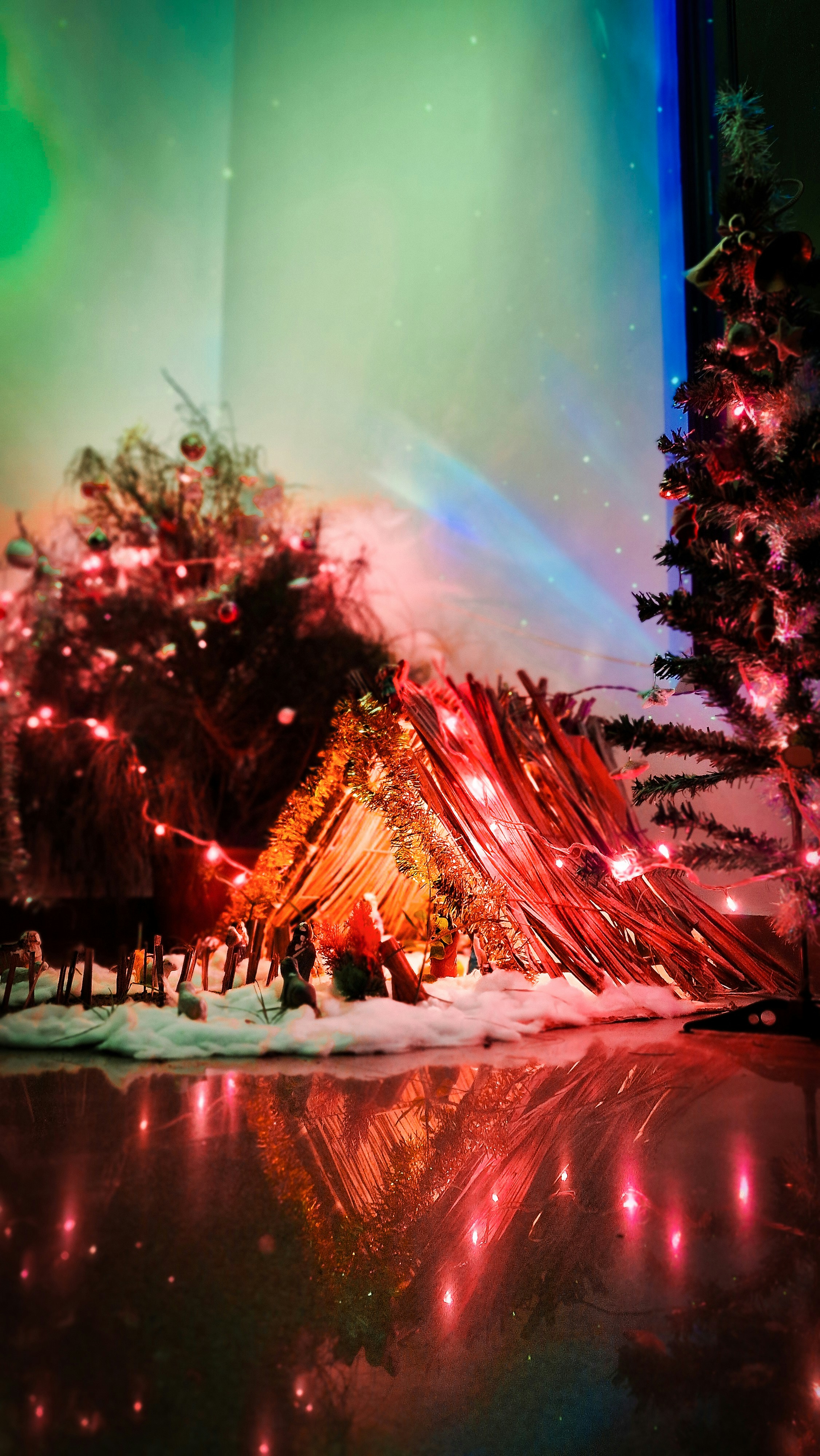 A cozy red-lit teepee village sits in snow beneath a colorful aurora backdrop, its lights mirrored on a glossy floor.