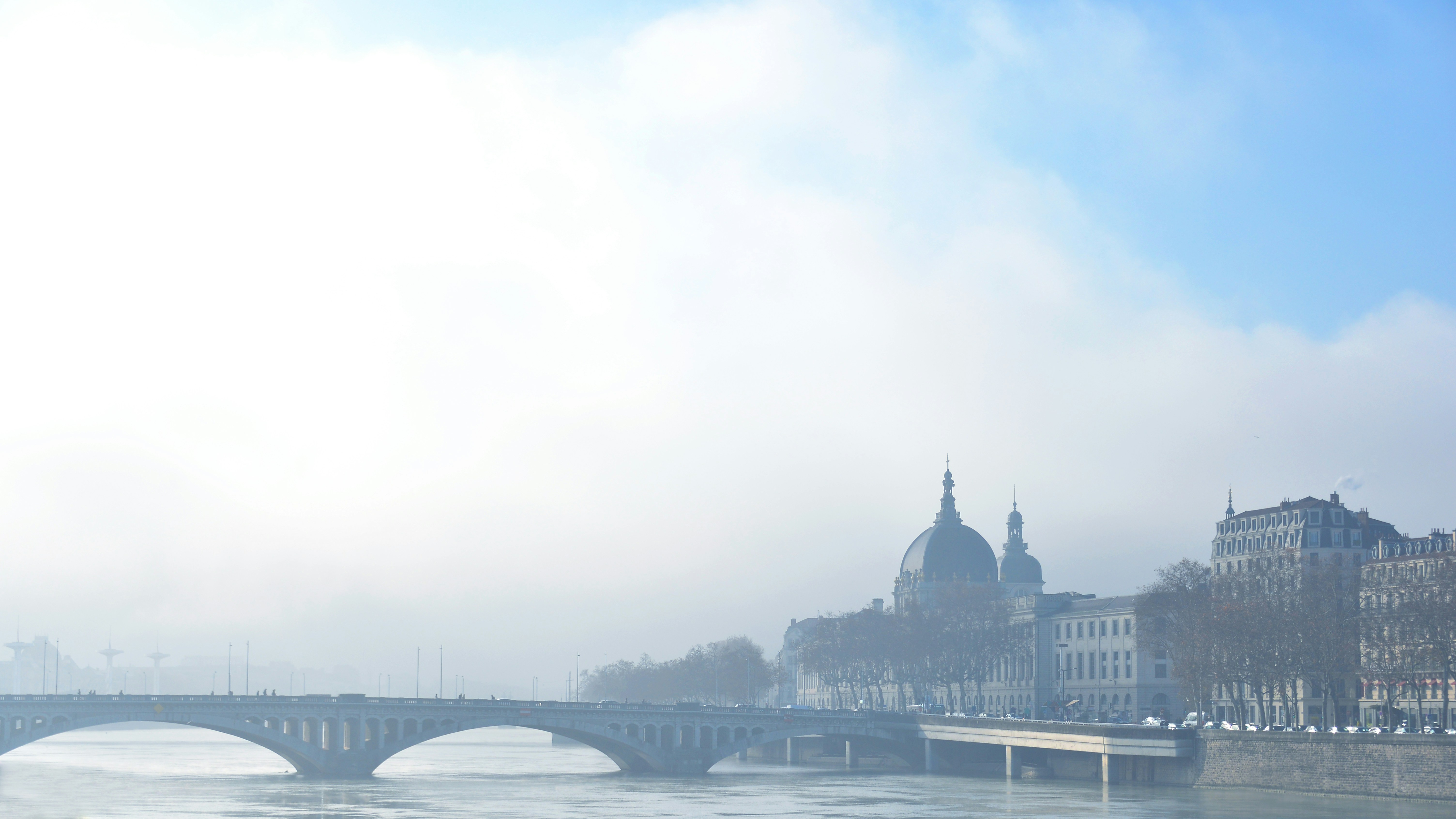 Bridge and distant buildings partially obscured by morning fog over a calm river.