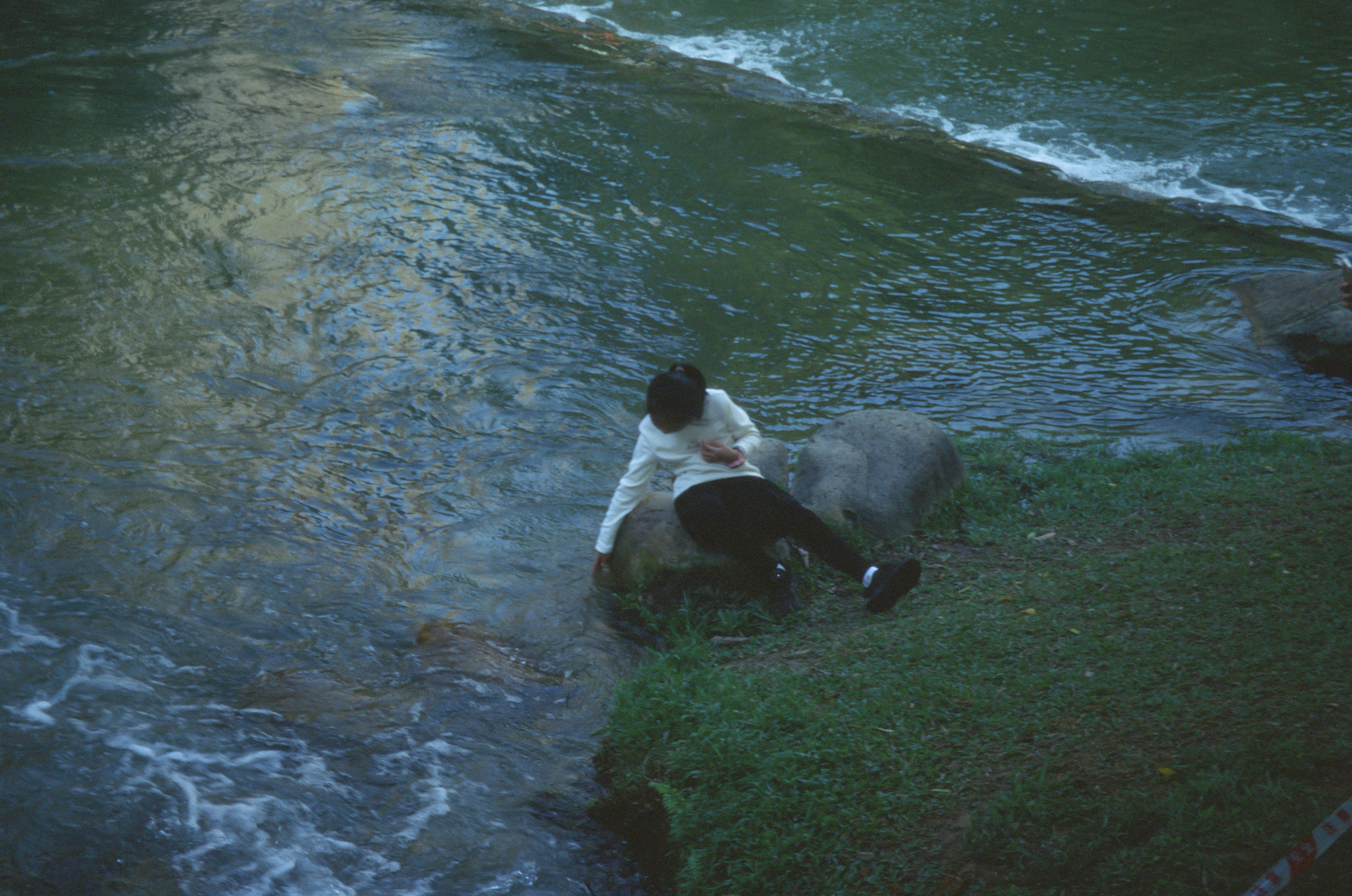 A person sits on a weathered rock at the water's edge, with rippling water and a grassy bank forming the backdrop.