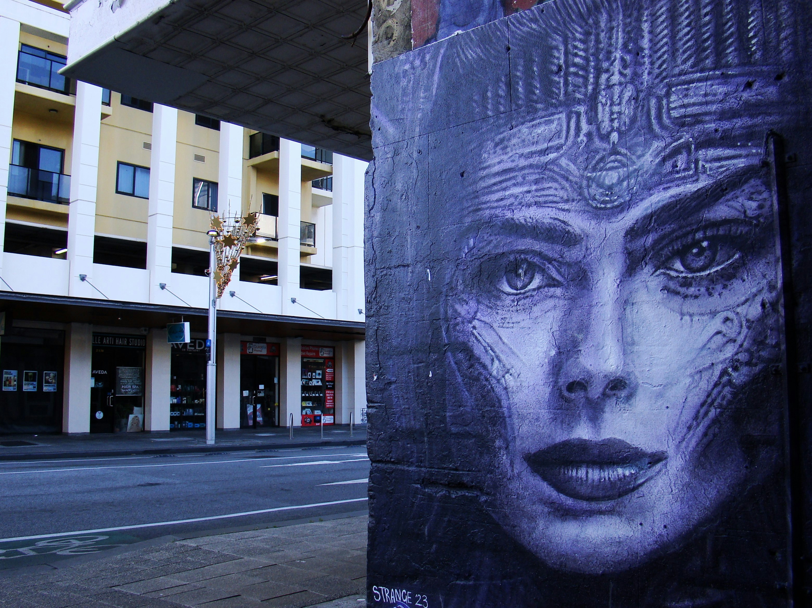 Blue-toned mural painting of a woman's face dominates a street scene, with storefronts and a modern building in the background.