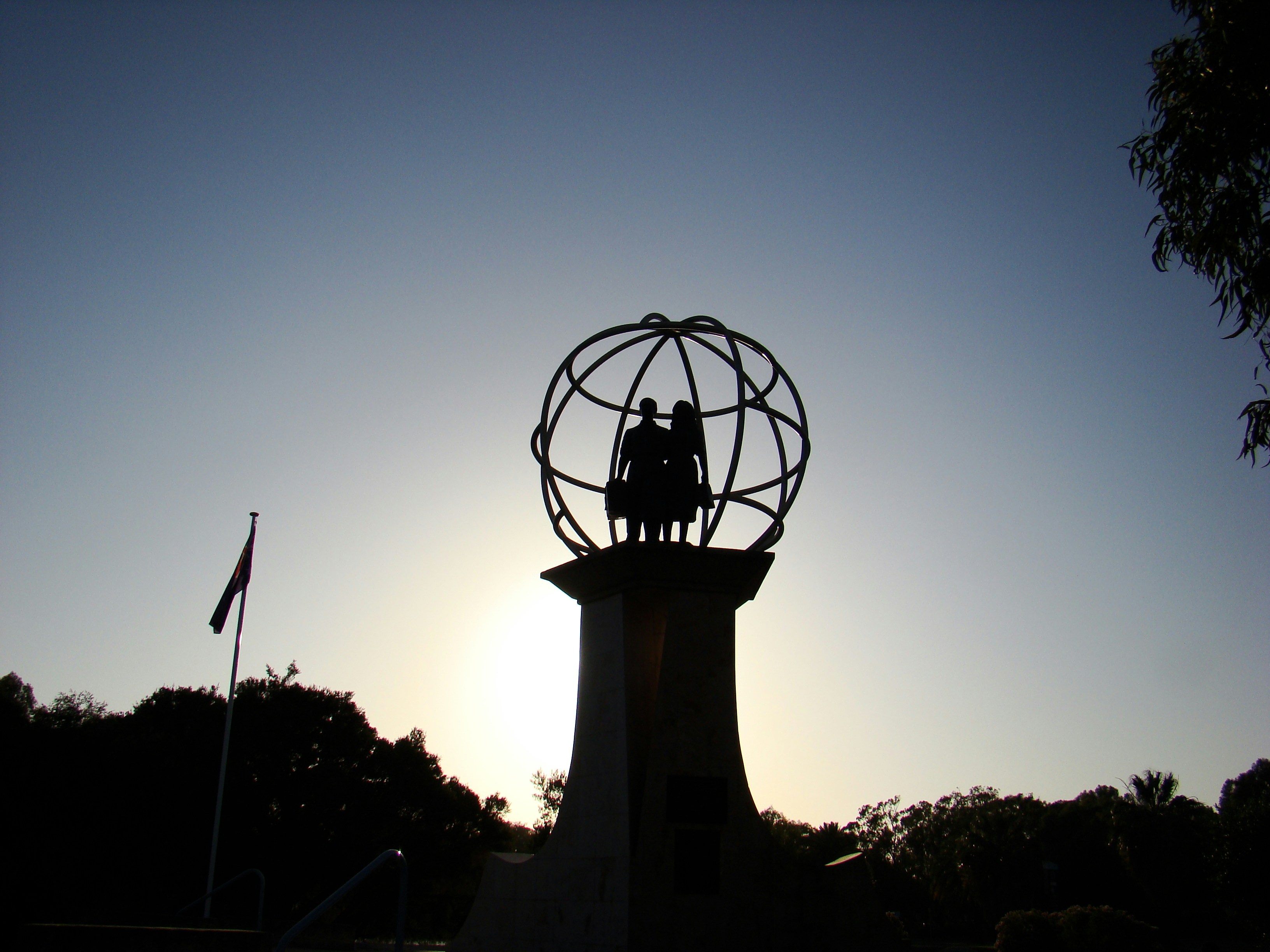 Silhouette of a globe sculpture atop a pedestal against a sunset, with the sun low on the horizon creating a halo and dark treeline in the foreground.