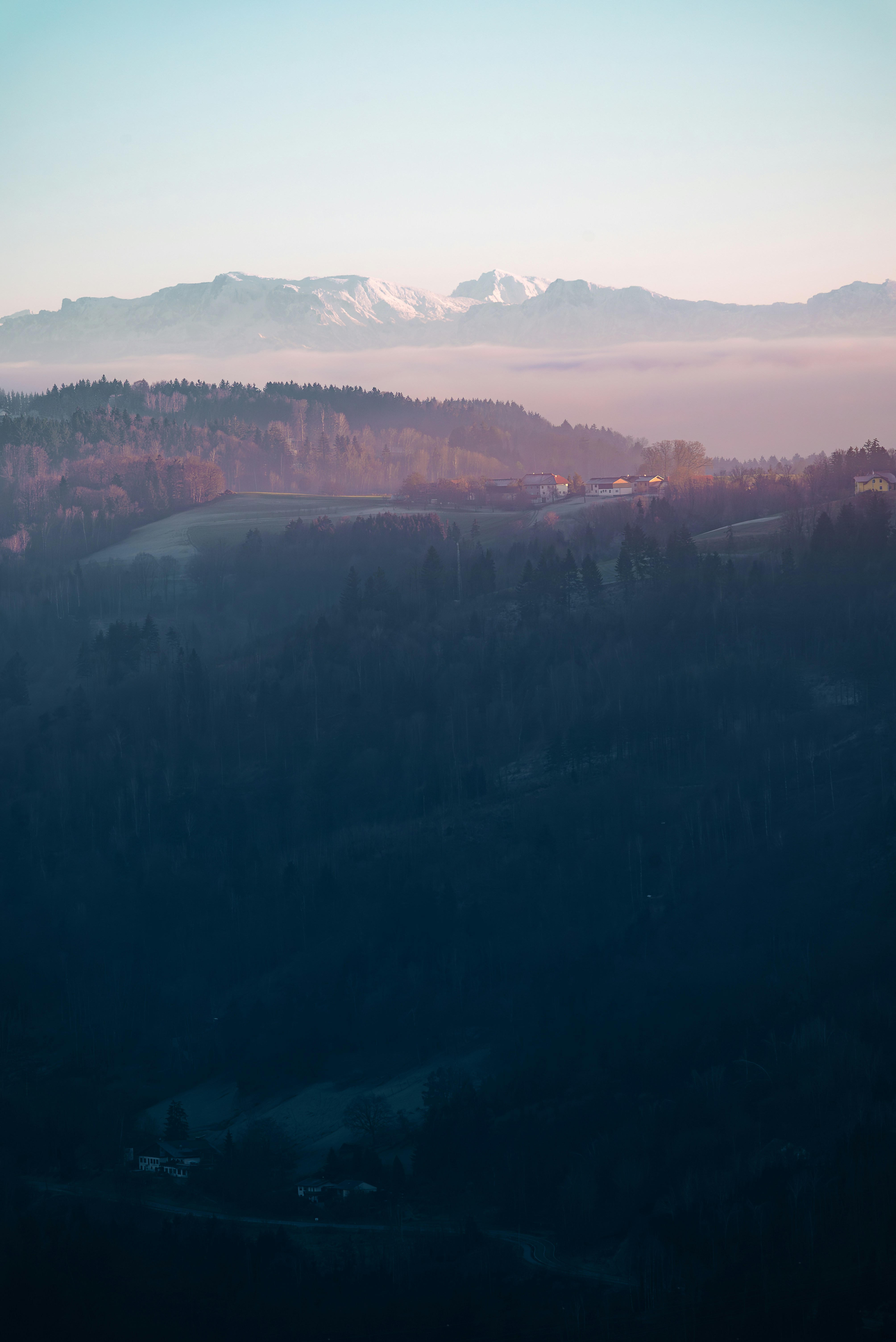 Snow-capped mountains rise above misty hills with scattered houses in soft morning light.