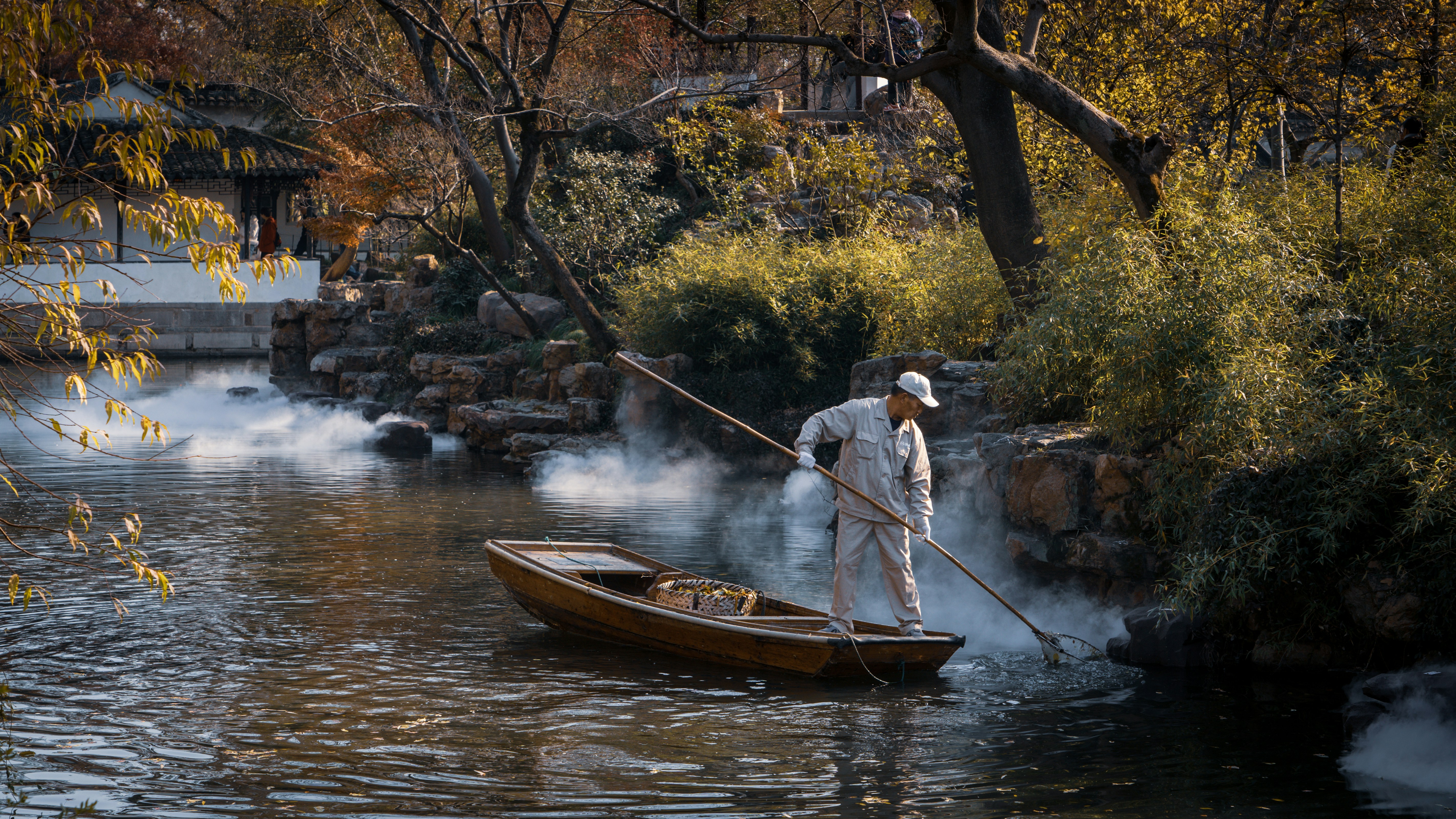 Person in white attire guides a small wooden boat along a serene river surrounded by autumn foliage.