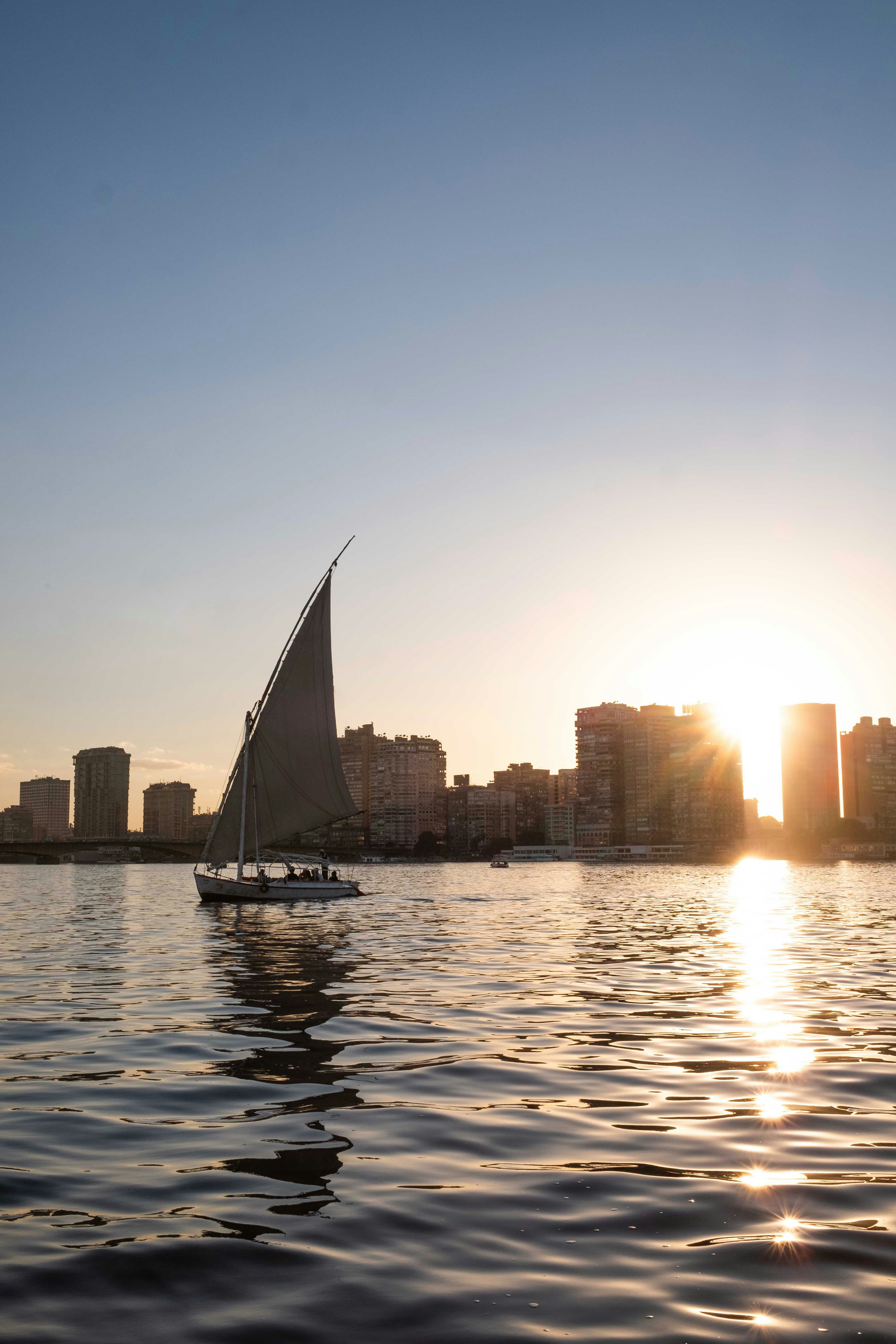 Sailboat gliding on rippling water with a city skyline silhouetted against a setting sun.
