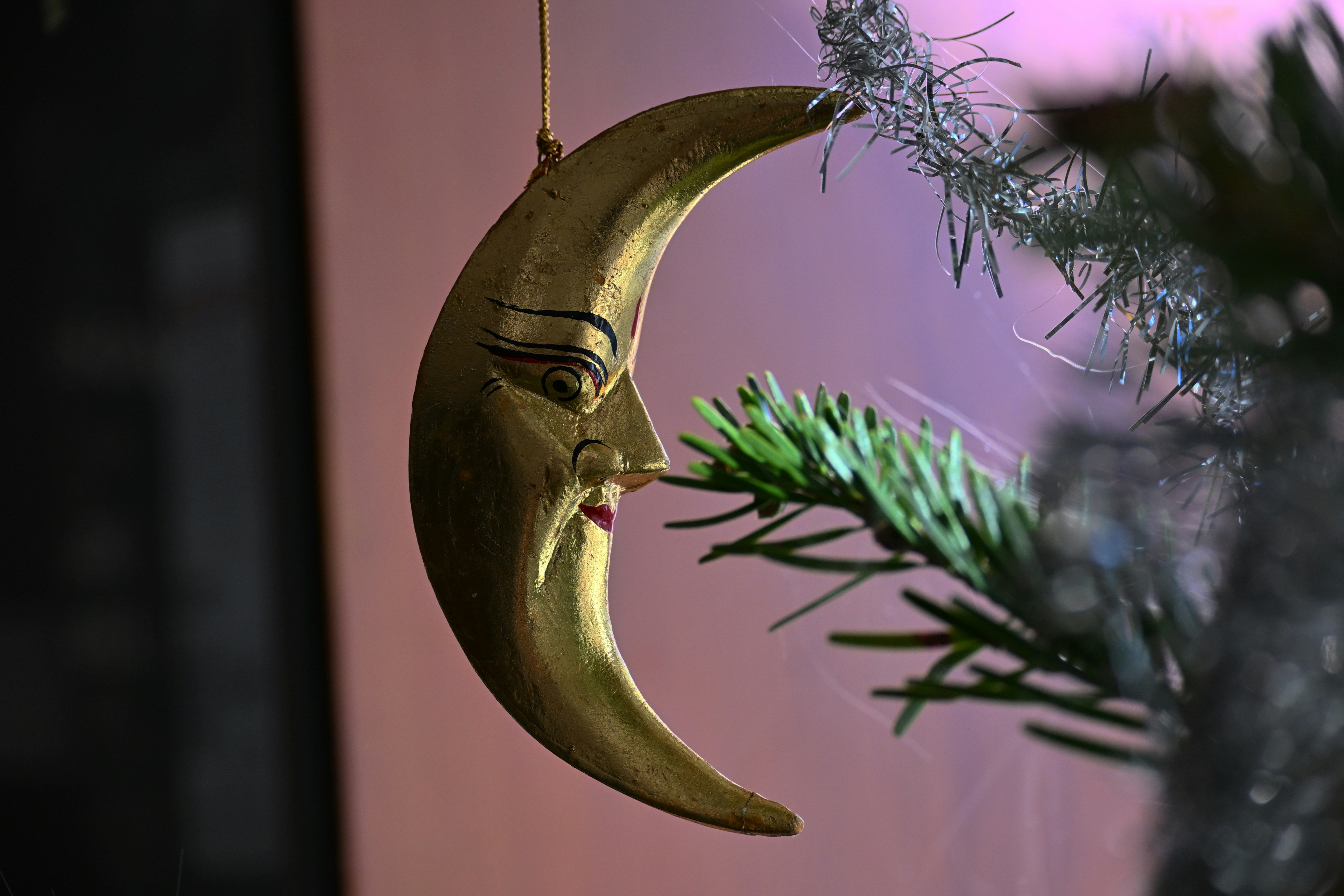 Golden crescent moon ornament with a painted face, hanging amidst evergreen branches. Soft light enhances its vibrant colors.