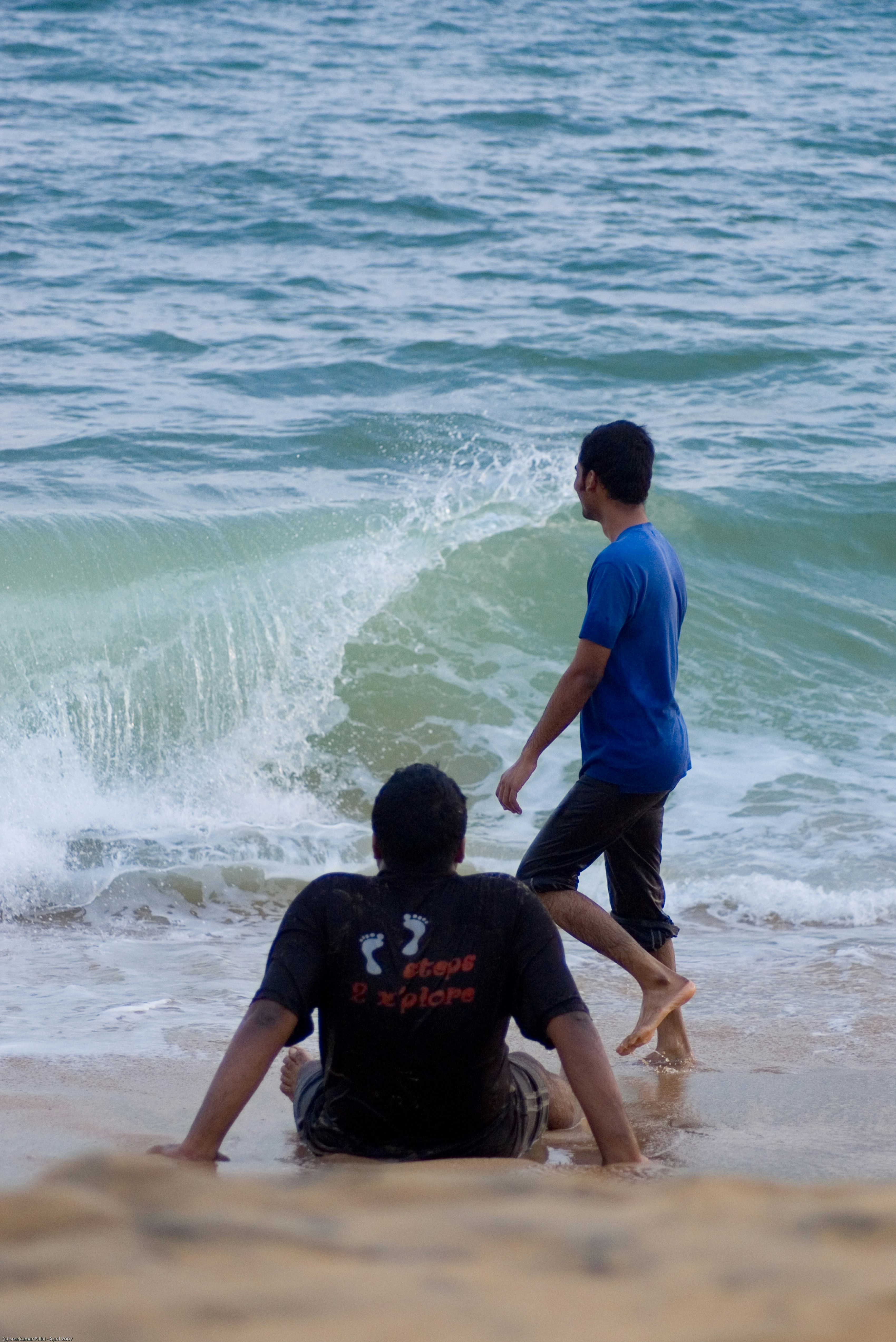 A photograph captures two men on a sandy beach with a turquoise wave breaking nearby. One sits in the foreground, the other stands closer to the water.